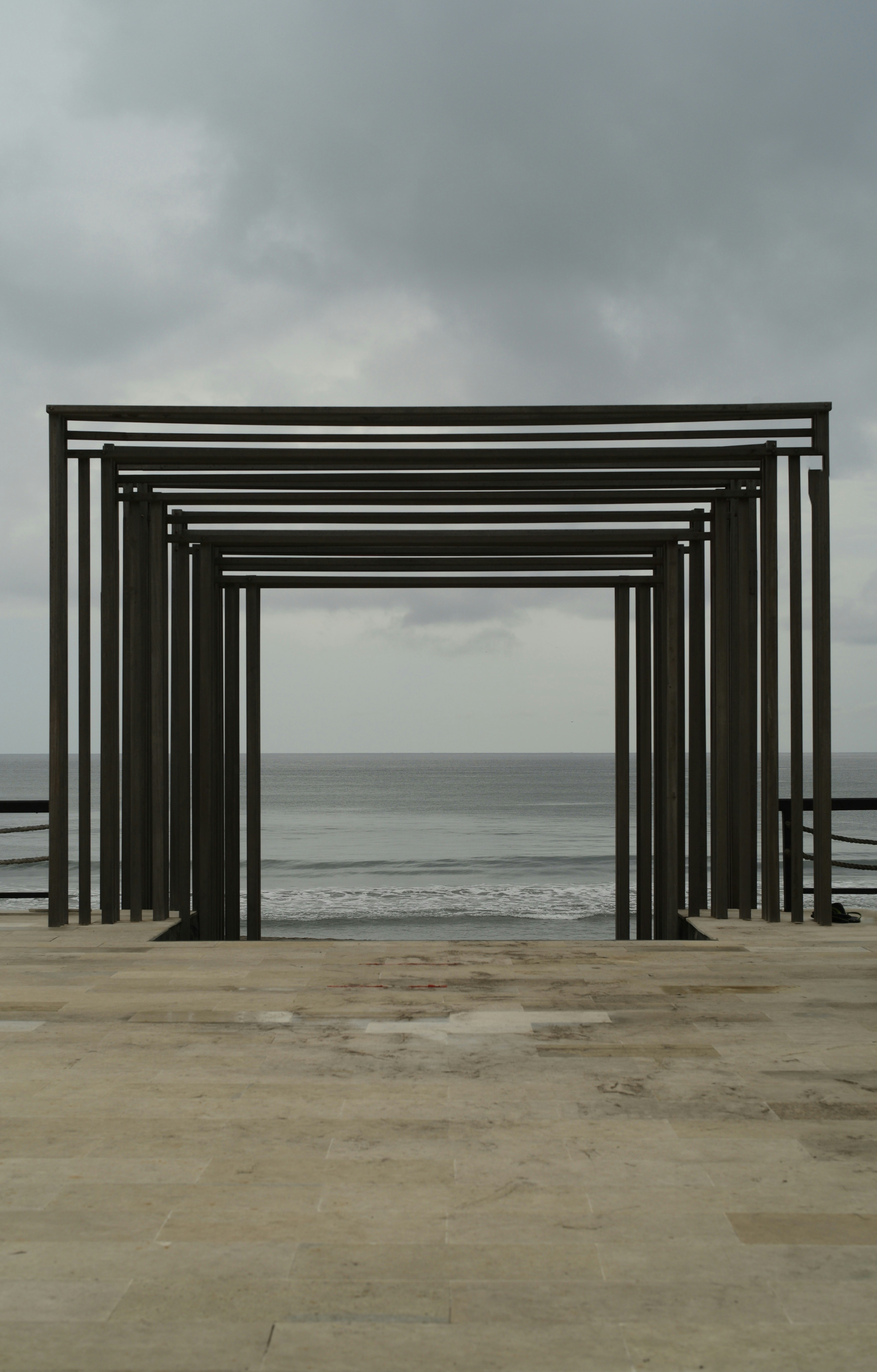 Rectangular steel frames form a tunnel toward the sea under an overcast sky.