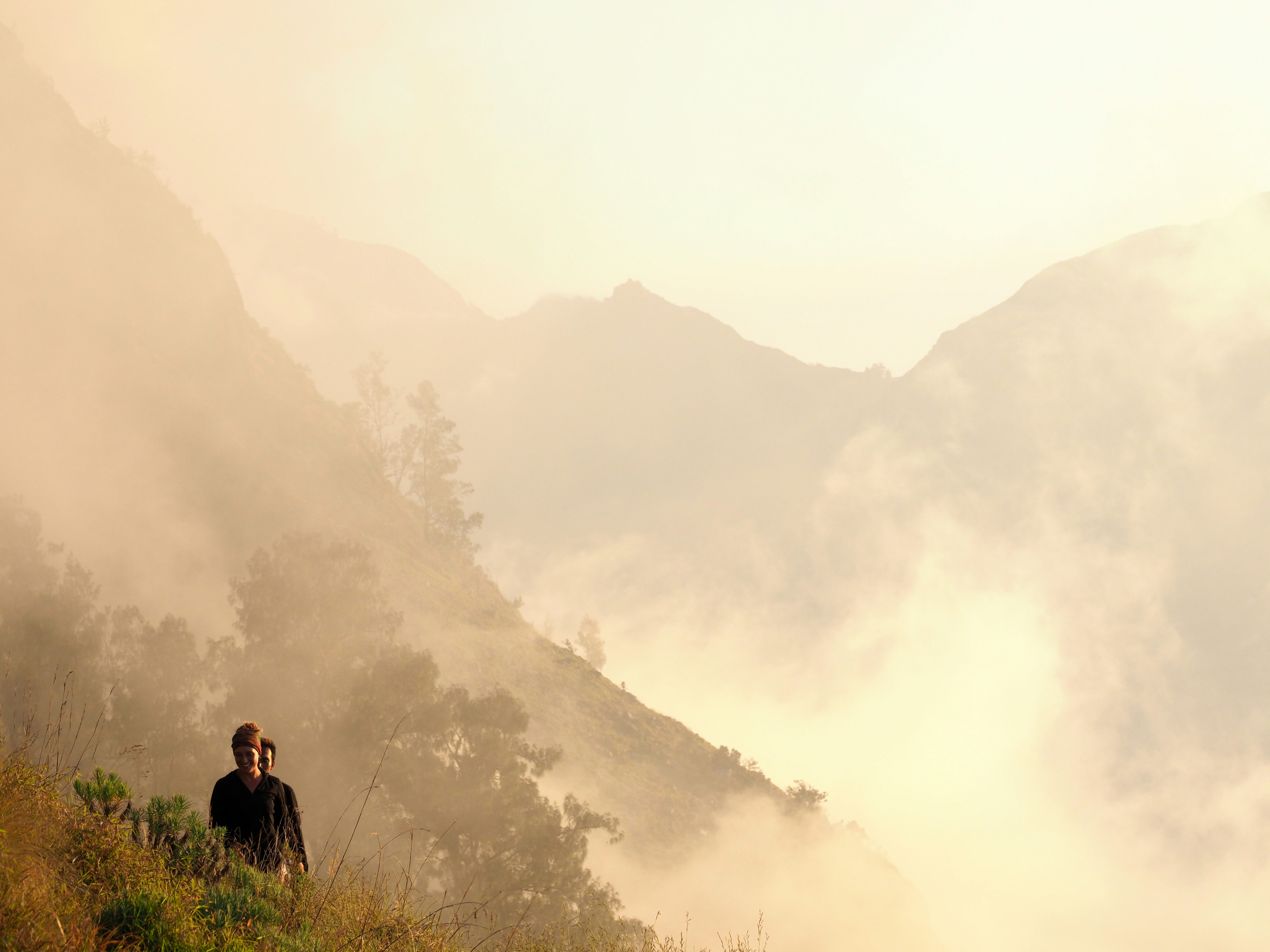 Hiker pauses on a misty mountain slope, enveloped by fog with rugged peaks in the background.
