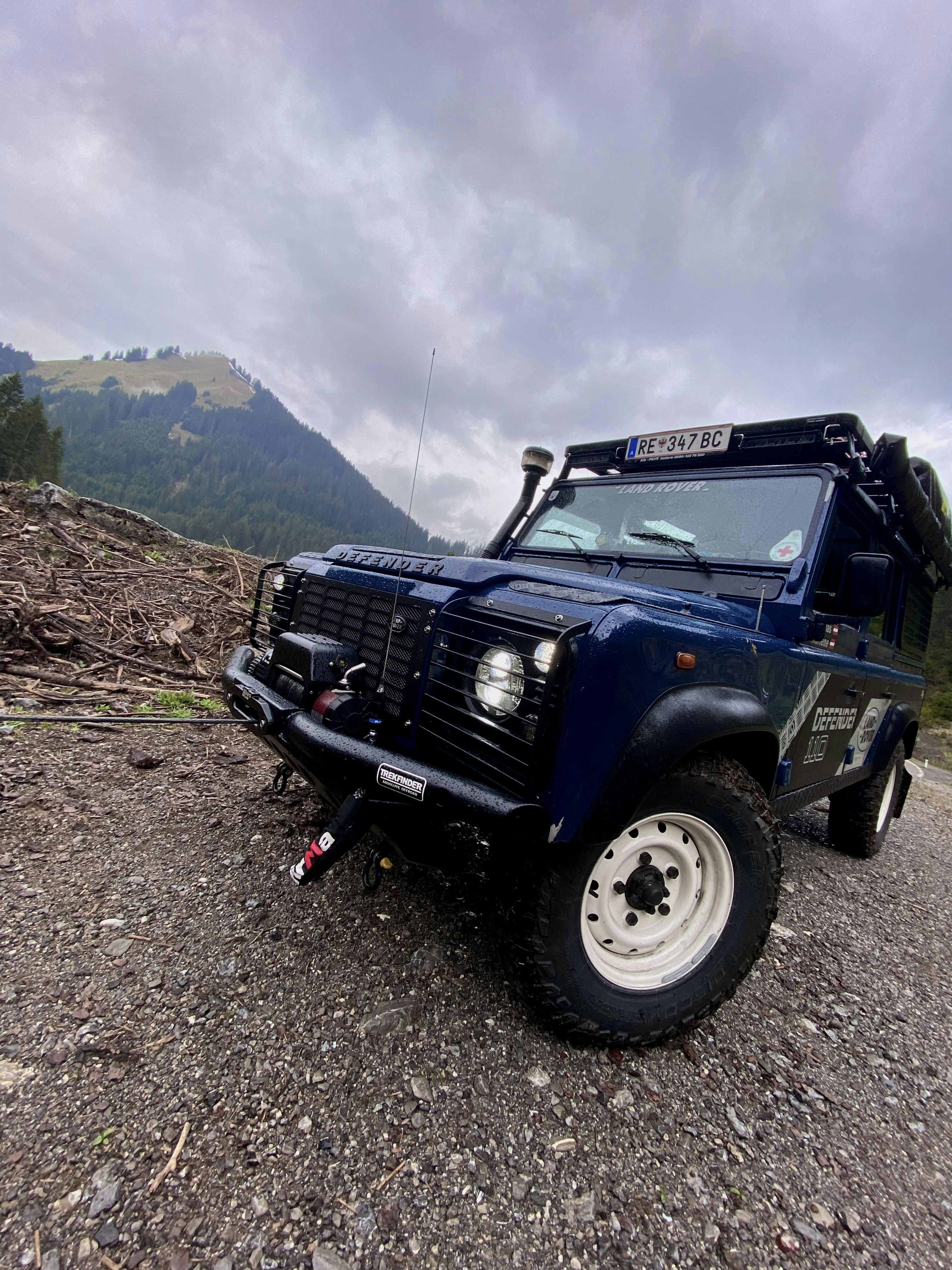 A blue Land Rover Defender parked on a gravel path, surrounded by lush mountains under a cloudy sky. The vehicle's rugged design highlights its off-road capabilities.