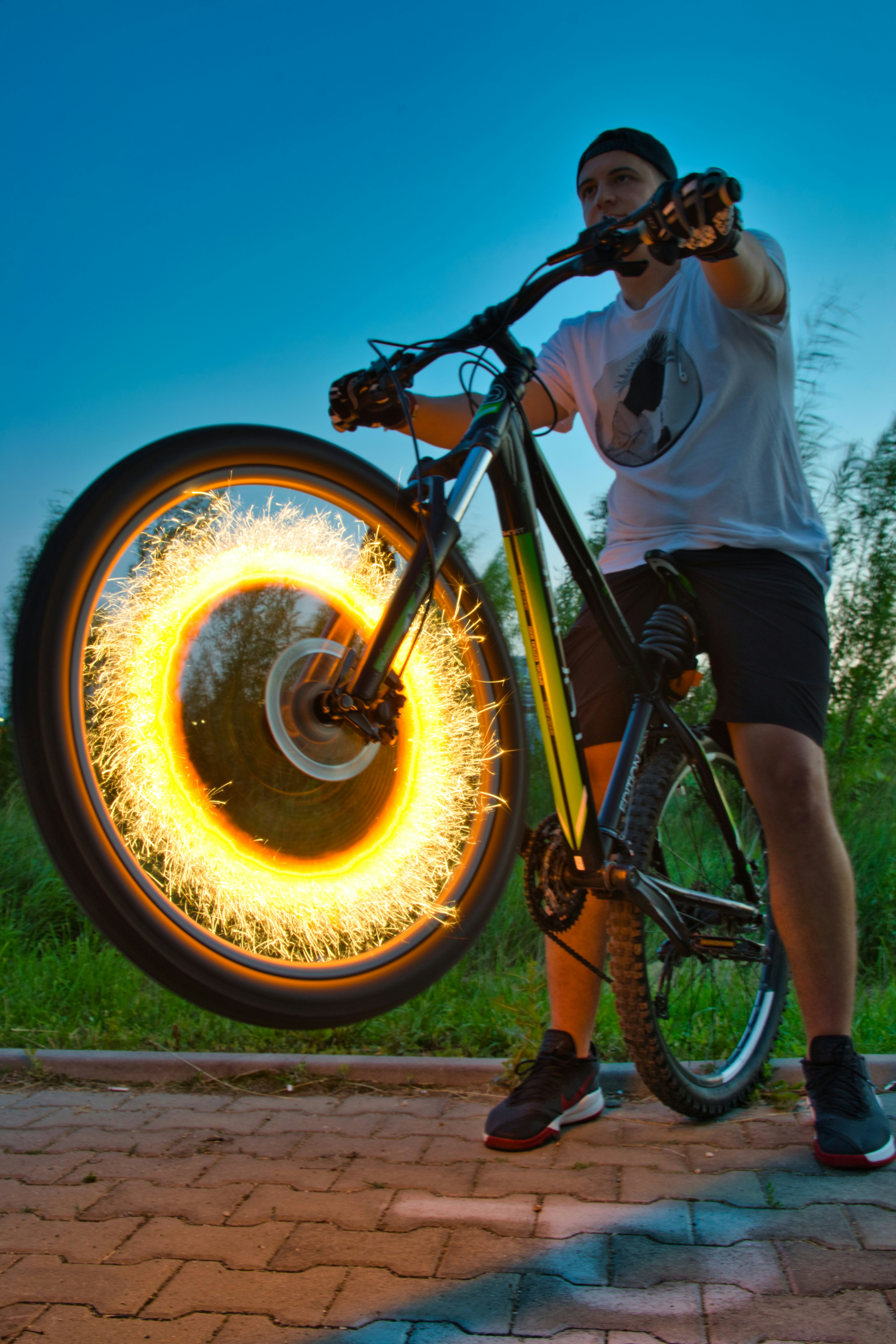 A cyclist performs a trick with a fiery wheel, illuminating the scene with vibrant sparks against a twilight backdrop.