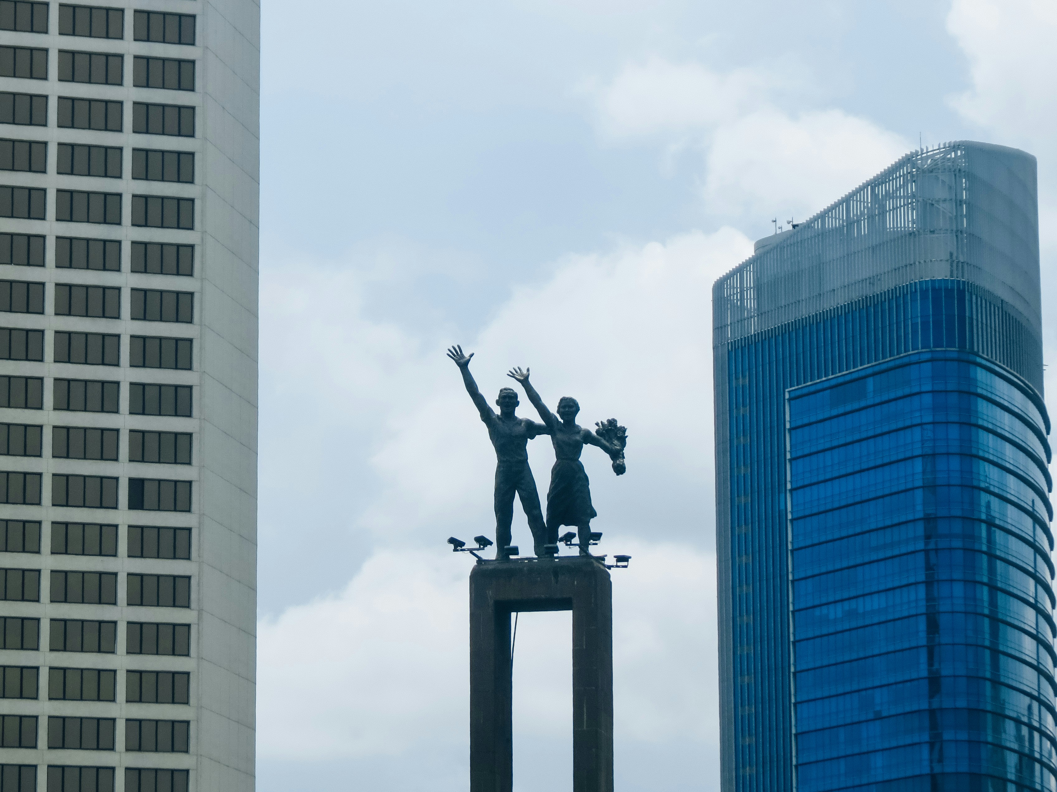 Sculpture of two figures celebrating atop a pedestal, surrounded by modern skyscrapers under a cloudy sky.