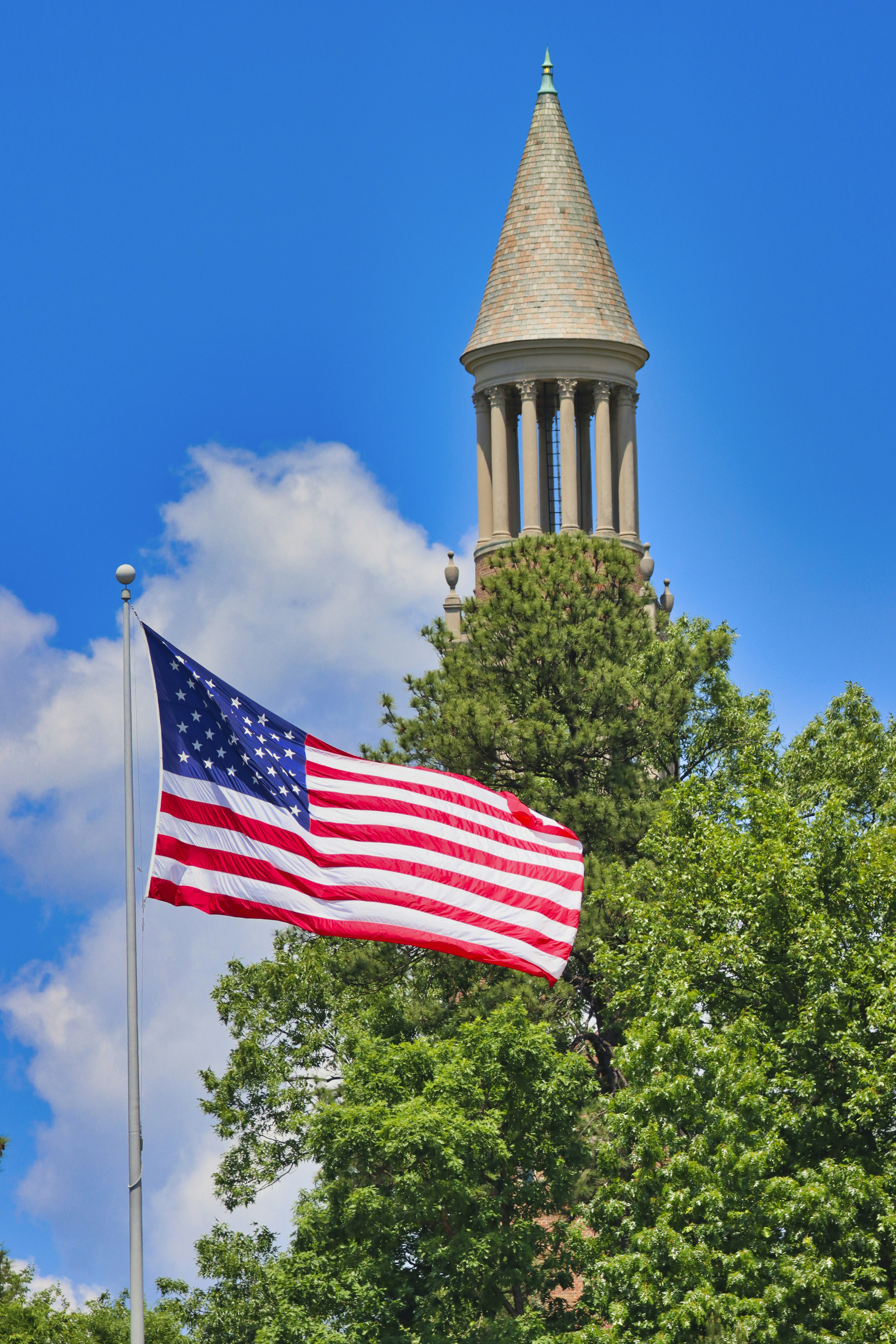 American flag fluttering in the breeze with a historic tower and lush greenery in the background.