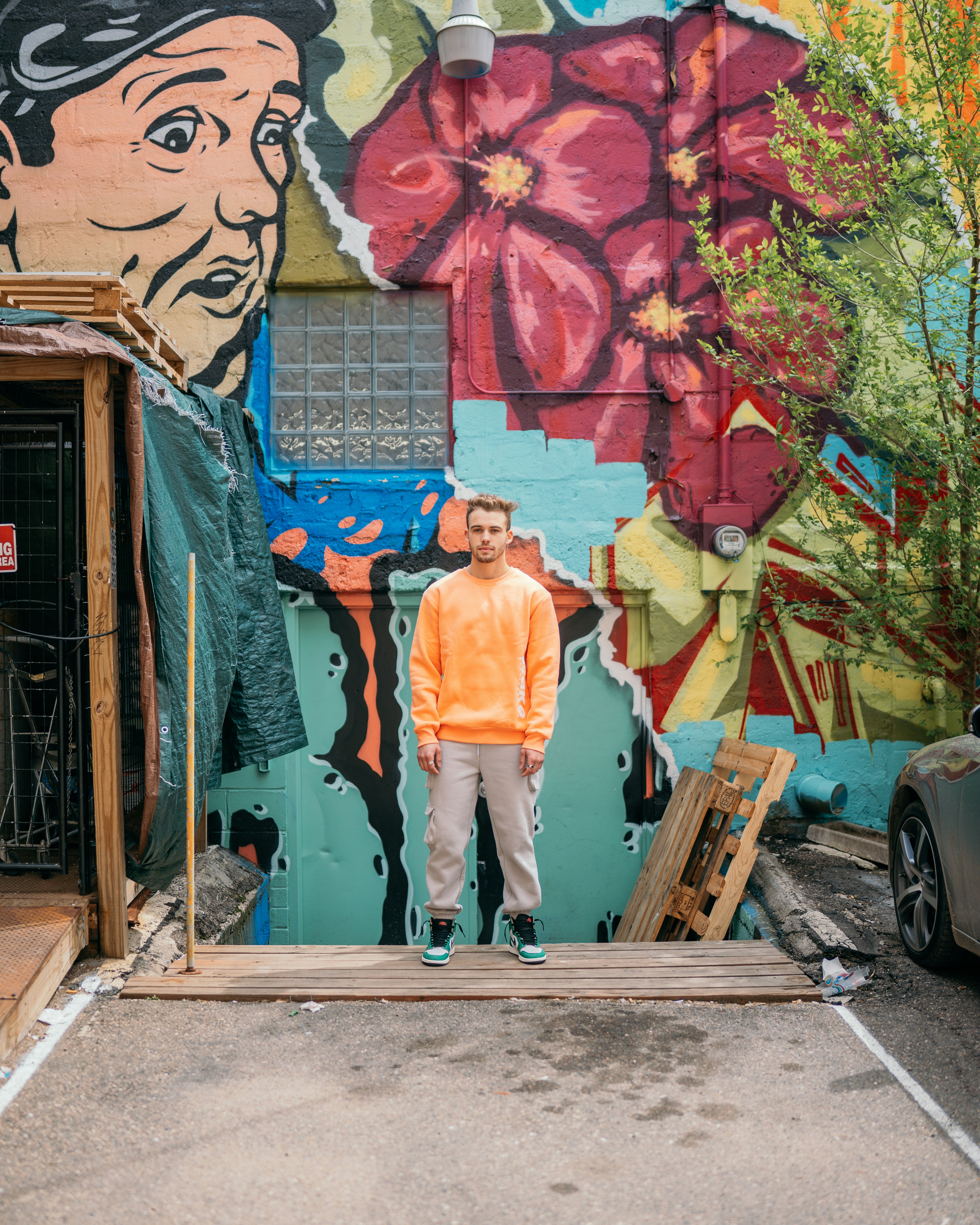 A young man stands confidently in front of a vibrant mural featuring a stylized face and floral elements, embodying the spirit of urban art.