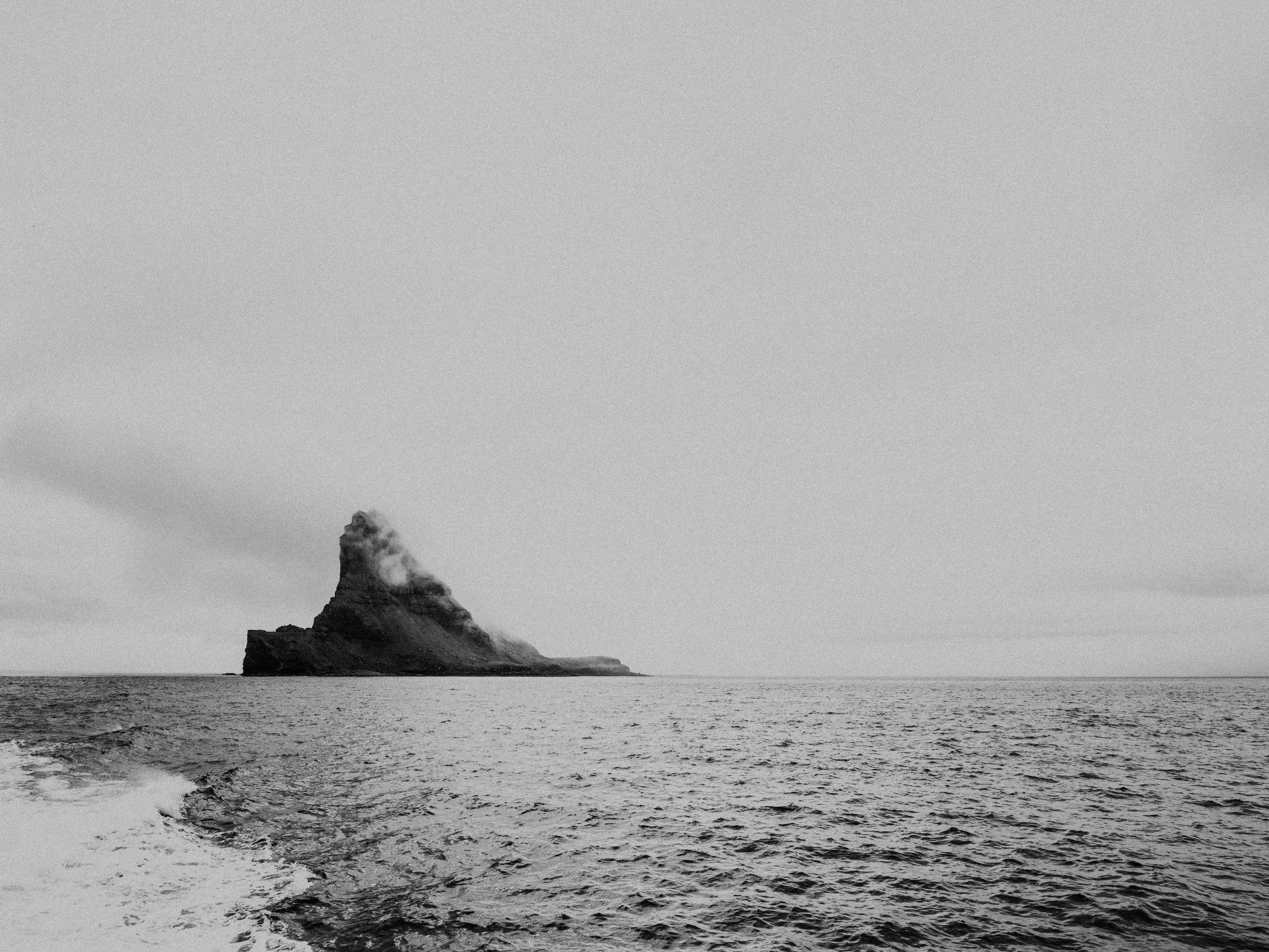 A solitary rock formation rises dramatically from the ocean, shrouded in mist and mystery. The monochromatic tones enhance the ethereal atmosphere.
