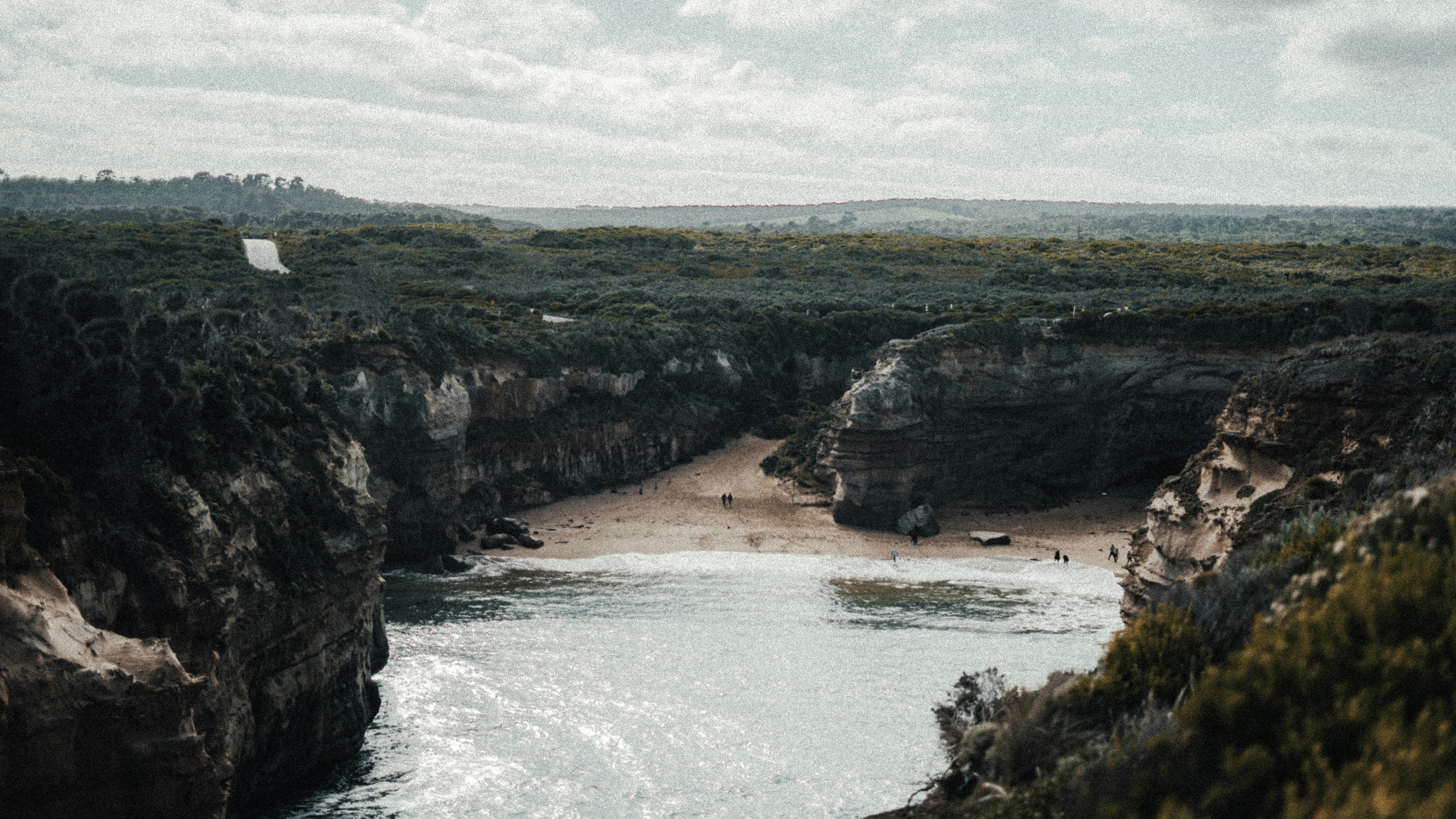 Rocky cliffs embrace a secluded beach with gentle waves under a cloudy sky.