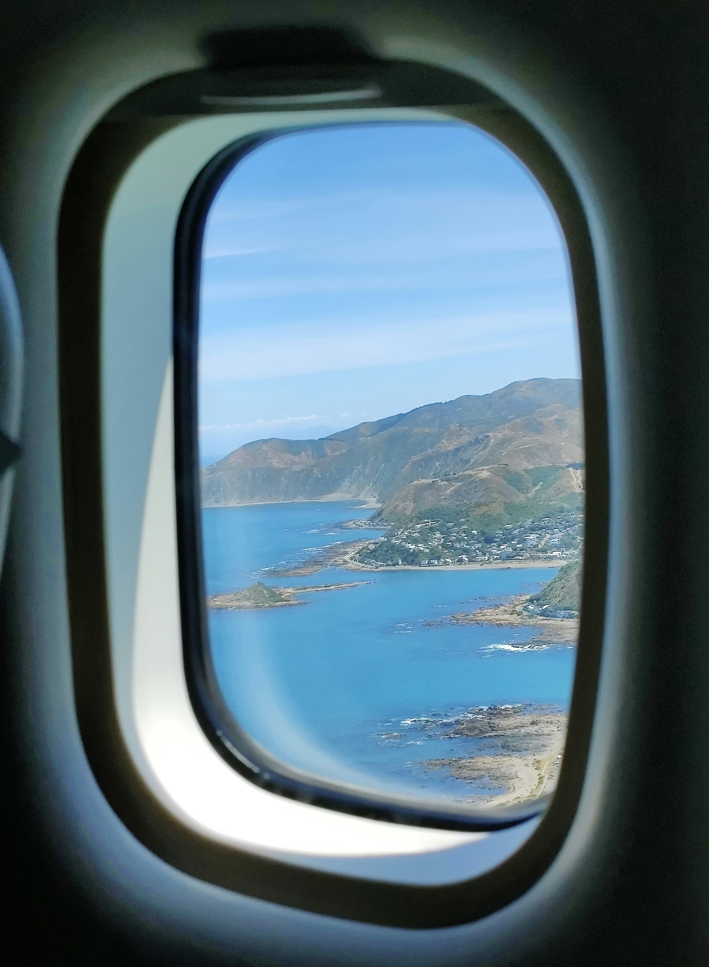 Photograph of a rugged coastline and blue sea framed by an airplane window, with distant hills and a small town along the shore.