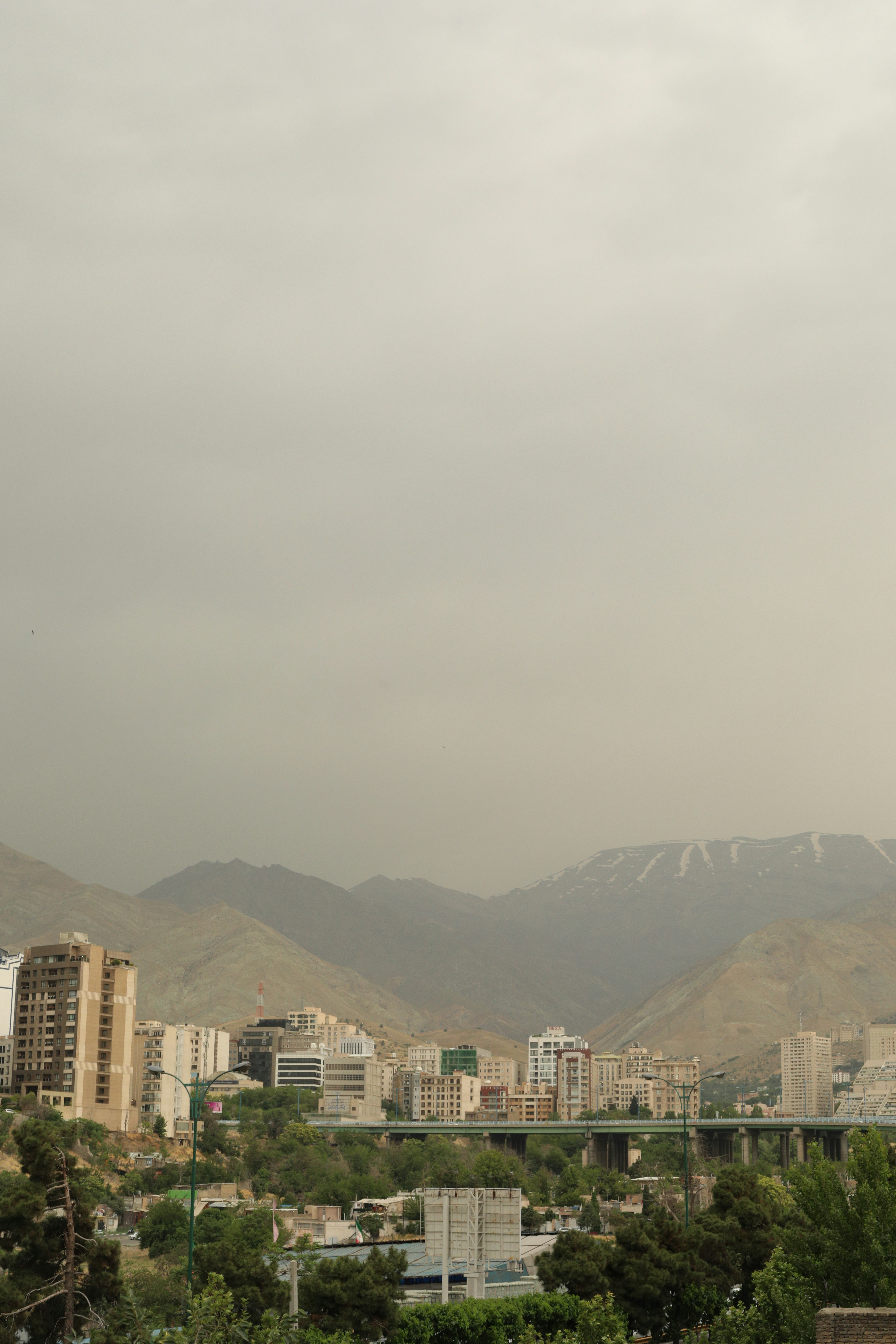 Cityscape featuring modern buildings juxtaposed with distant snow-capped mountains under a cloudy sky.