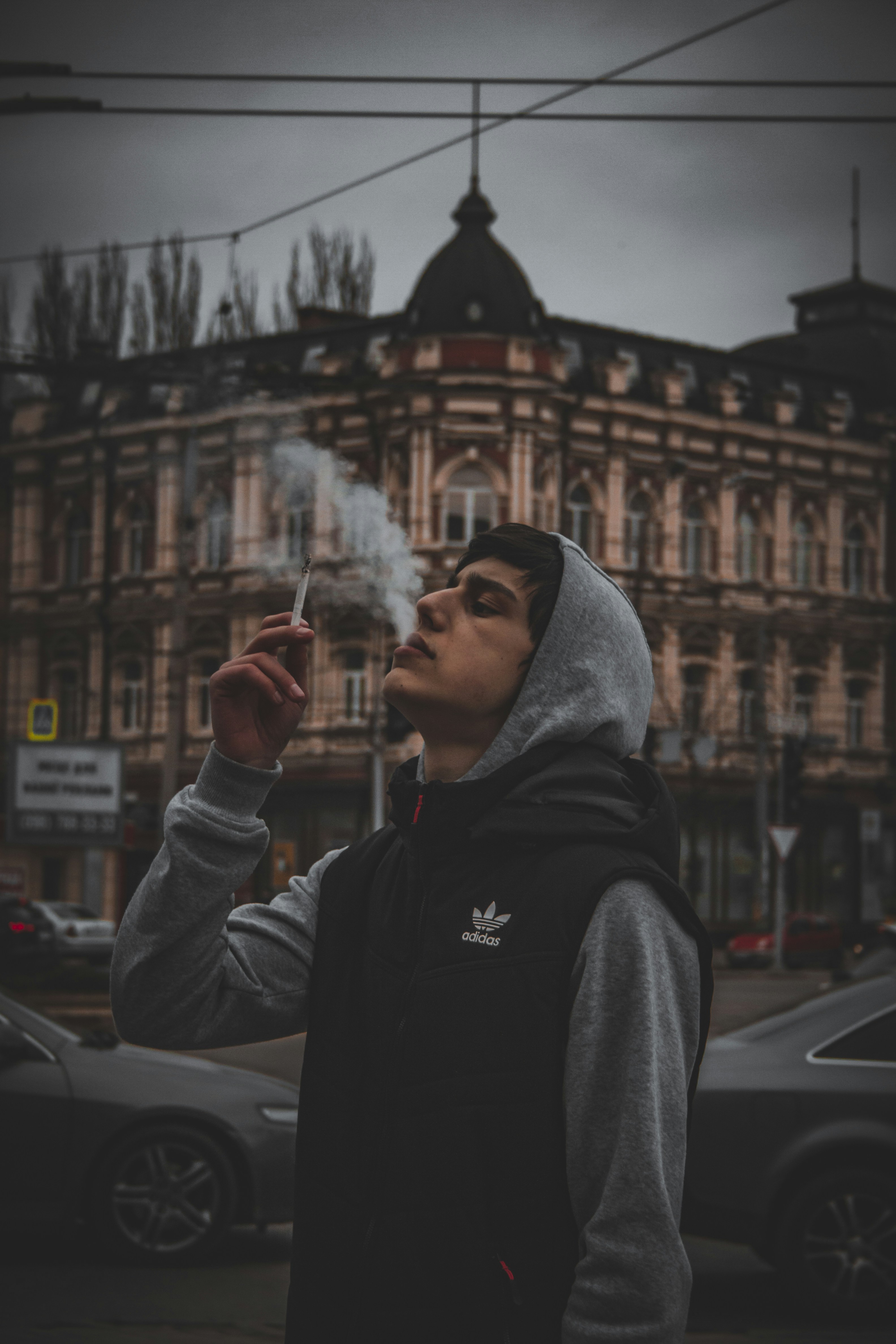 Young man smoking a cigarette in an urban setting, with a vintage building in the background. The scene captures a moment of contemplation amidst city life.