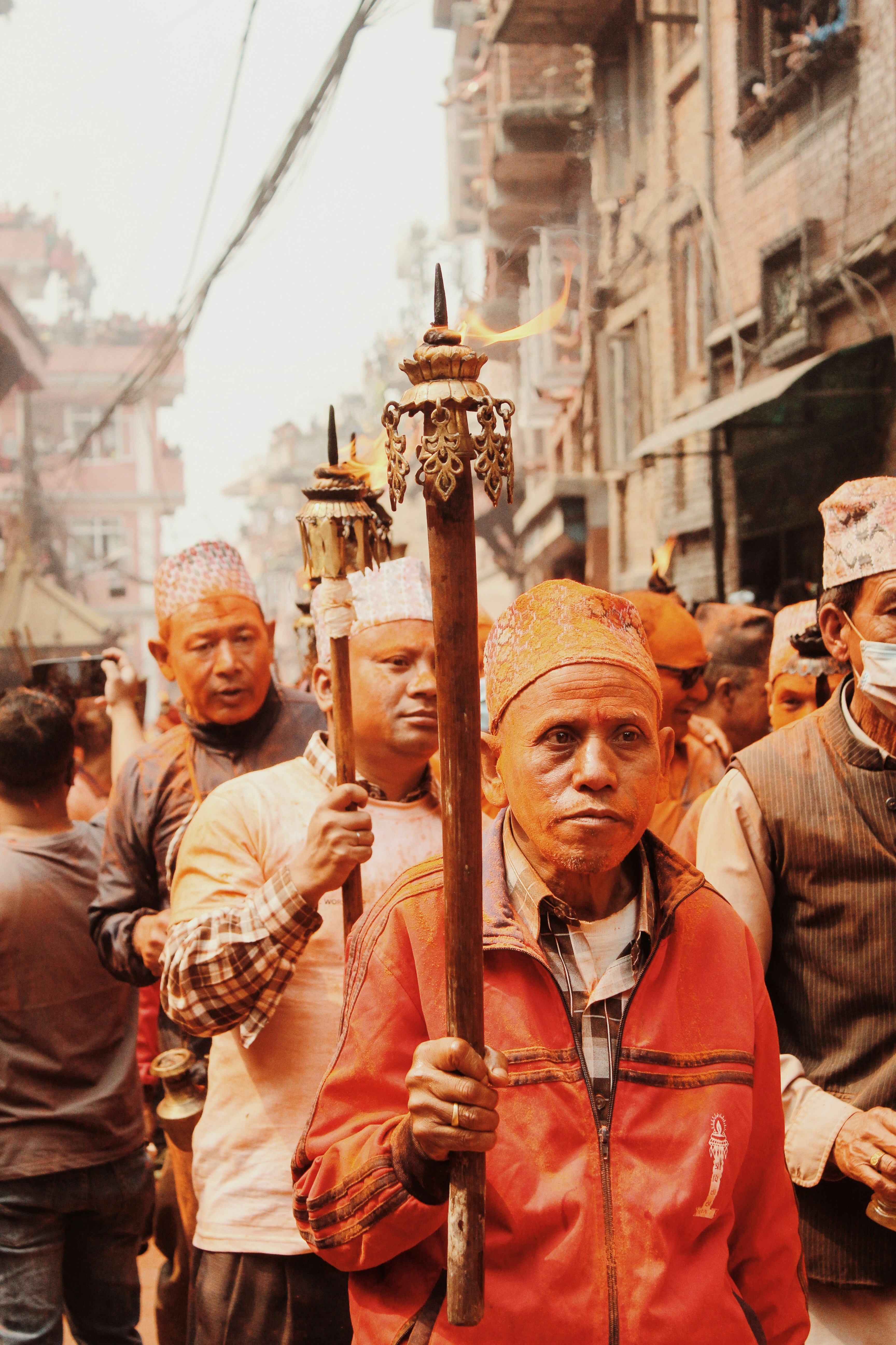 Men in traditional attire participate in a vibrant procession, holding torches amidst a bustling street scene. The atmosphere reflects cultural significance and community spirit.