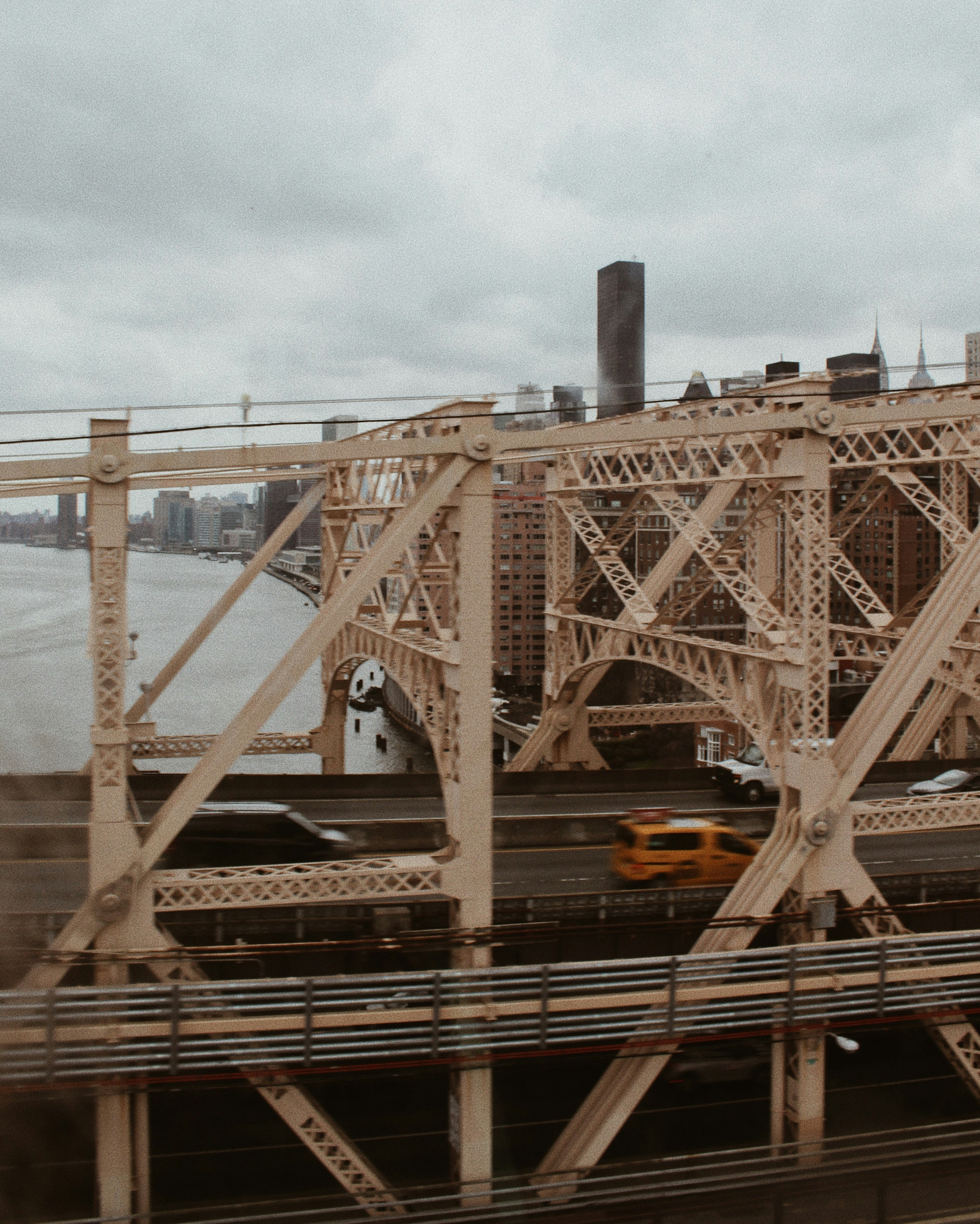 Overcast skyline behind rust-colored steel trusses of a busy bridge, with blurred traffic streaking along the roadway below. Photograph captures urban infrastructure and motion.