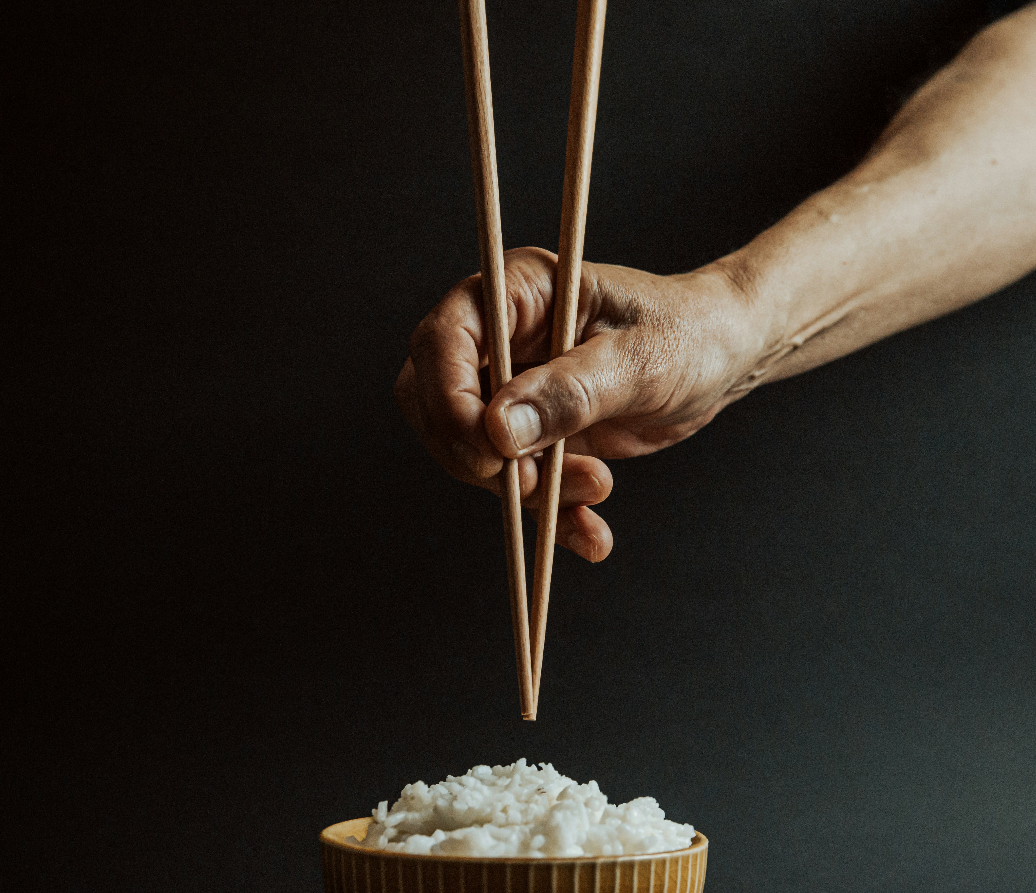 Silent Grip: Chopsticks Hover Over a Bowl of Rice Against a Dark Backdrop