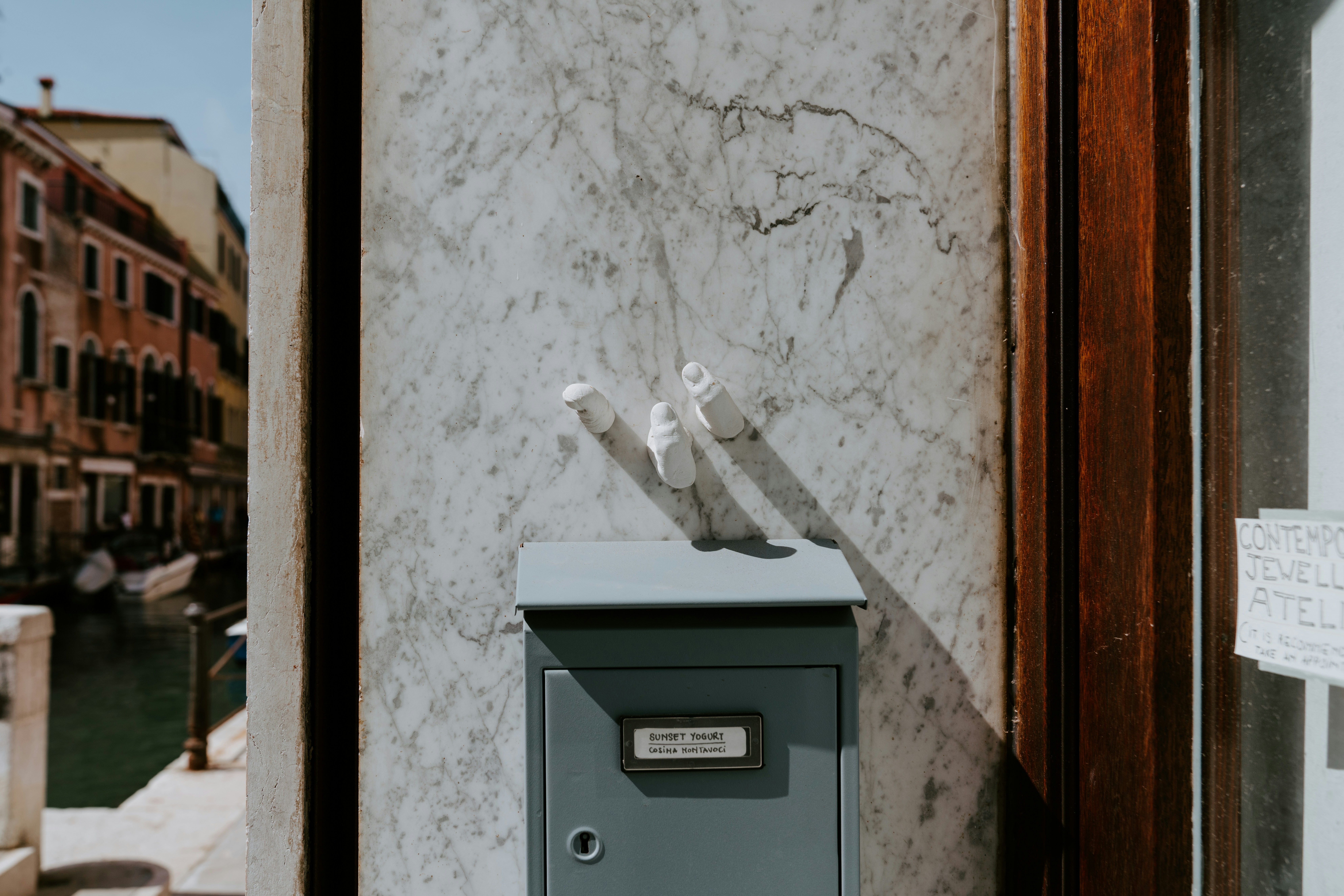 A weathered mail slot mounted on a marble wall, flanked by three white pipes, captures the essence of urban life against a serene canal backdrop.