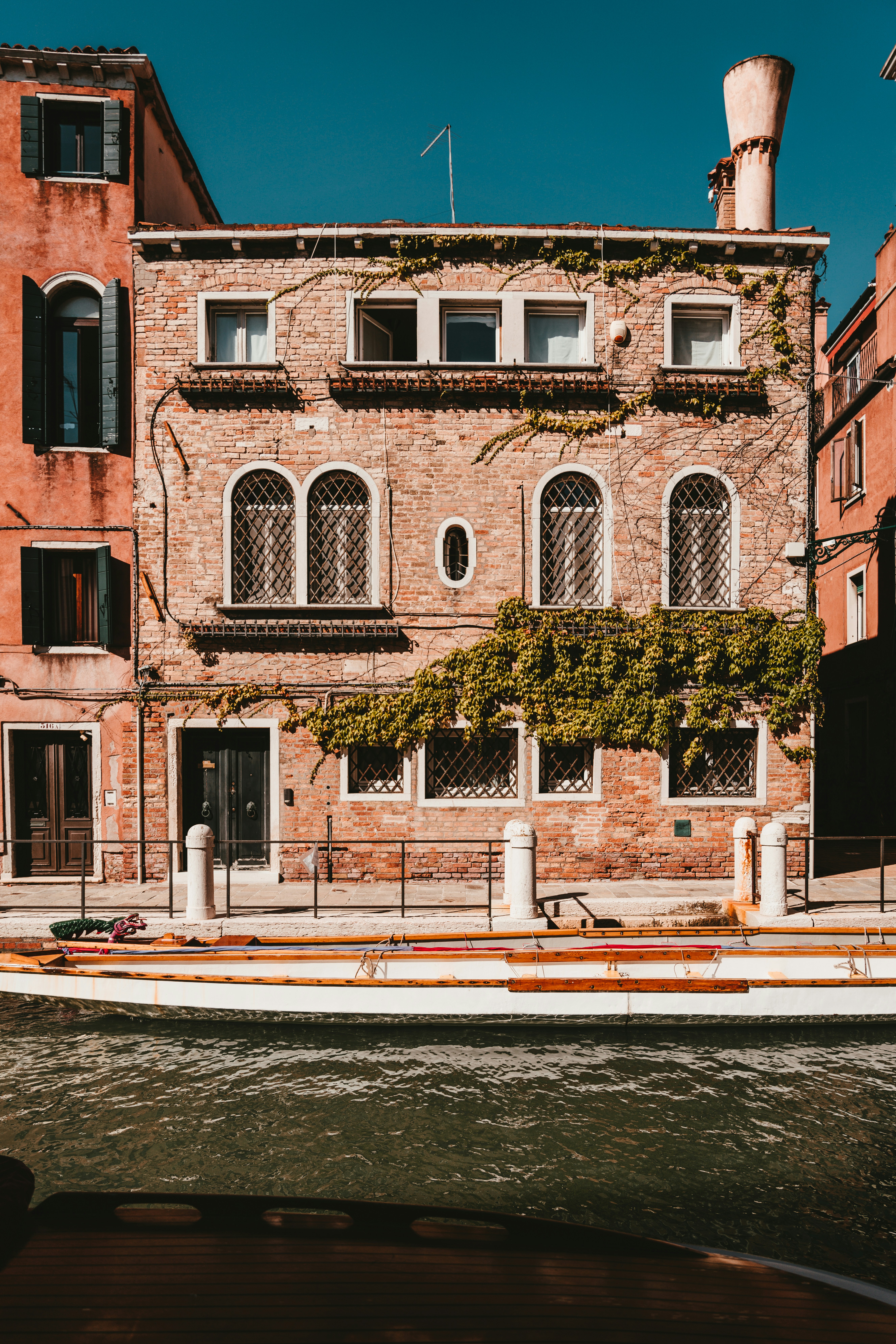 Historic Venetian building draped in ivy beside a tranquil canal, showcasing intricate window designs and vibrant colors. A gondola rests along the water's edge.