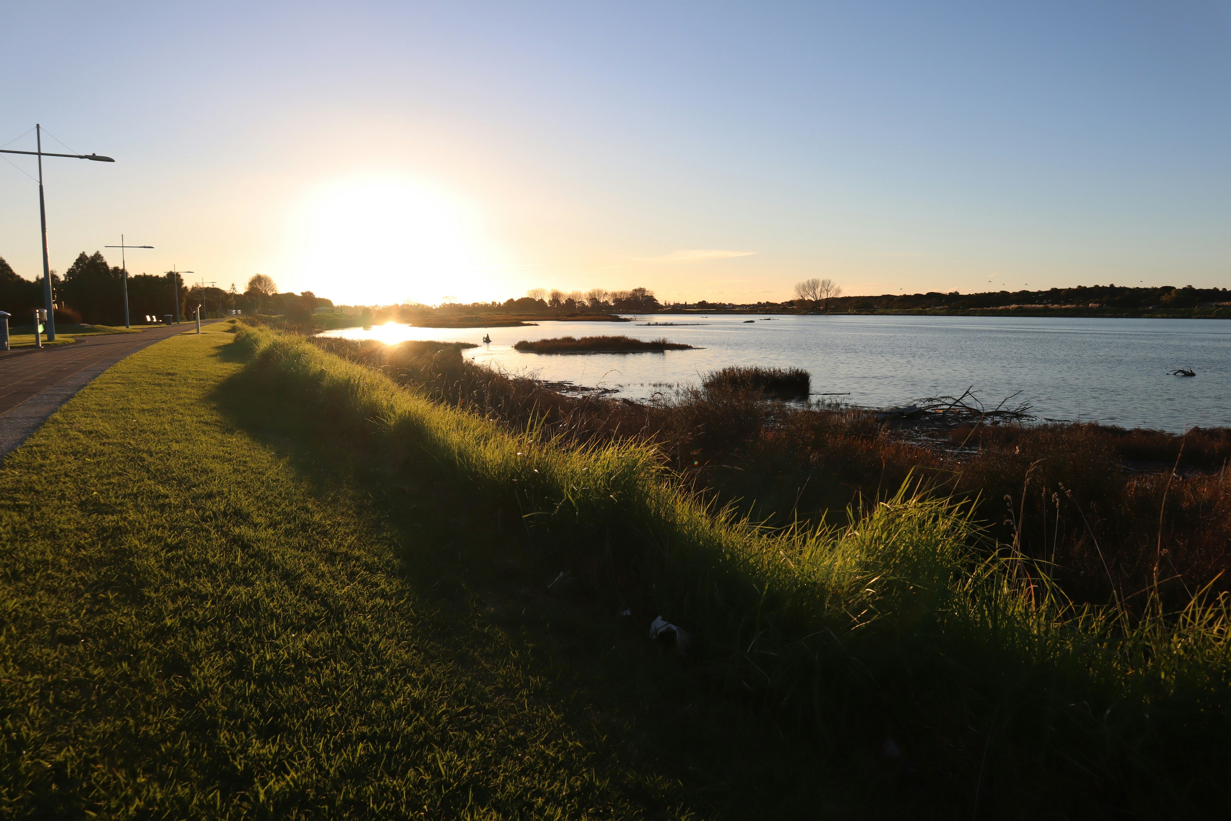 Sunset casting long shadows over a grassy riverbank with calm waters reflecting the warm glow.