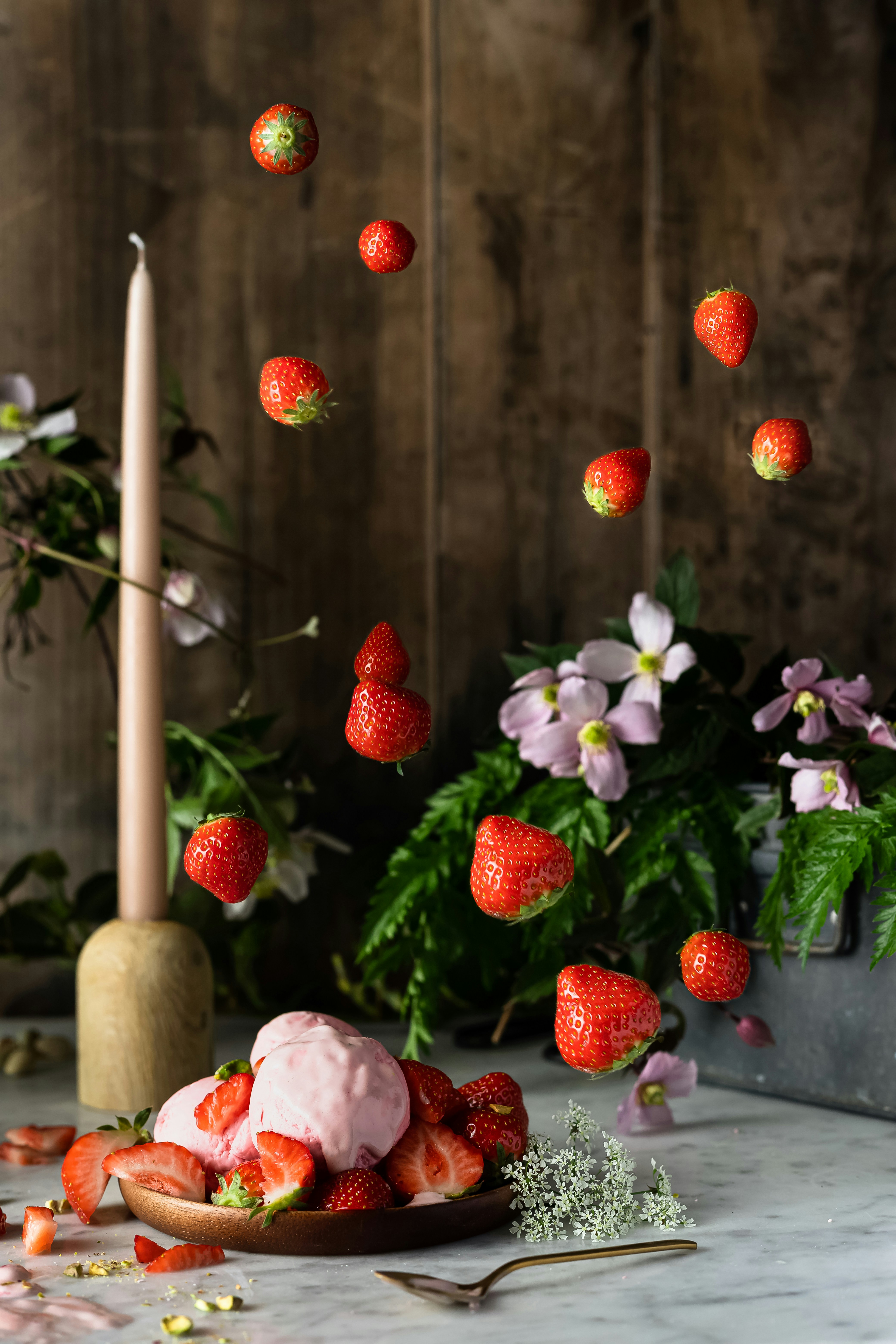Ripe strawberries levitate above a plate of strawberry ice cream on a marble surface with floral decor.