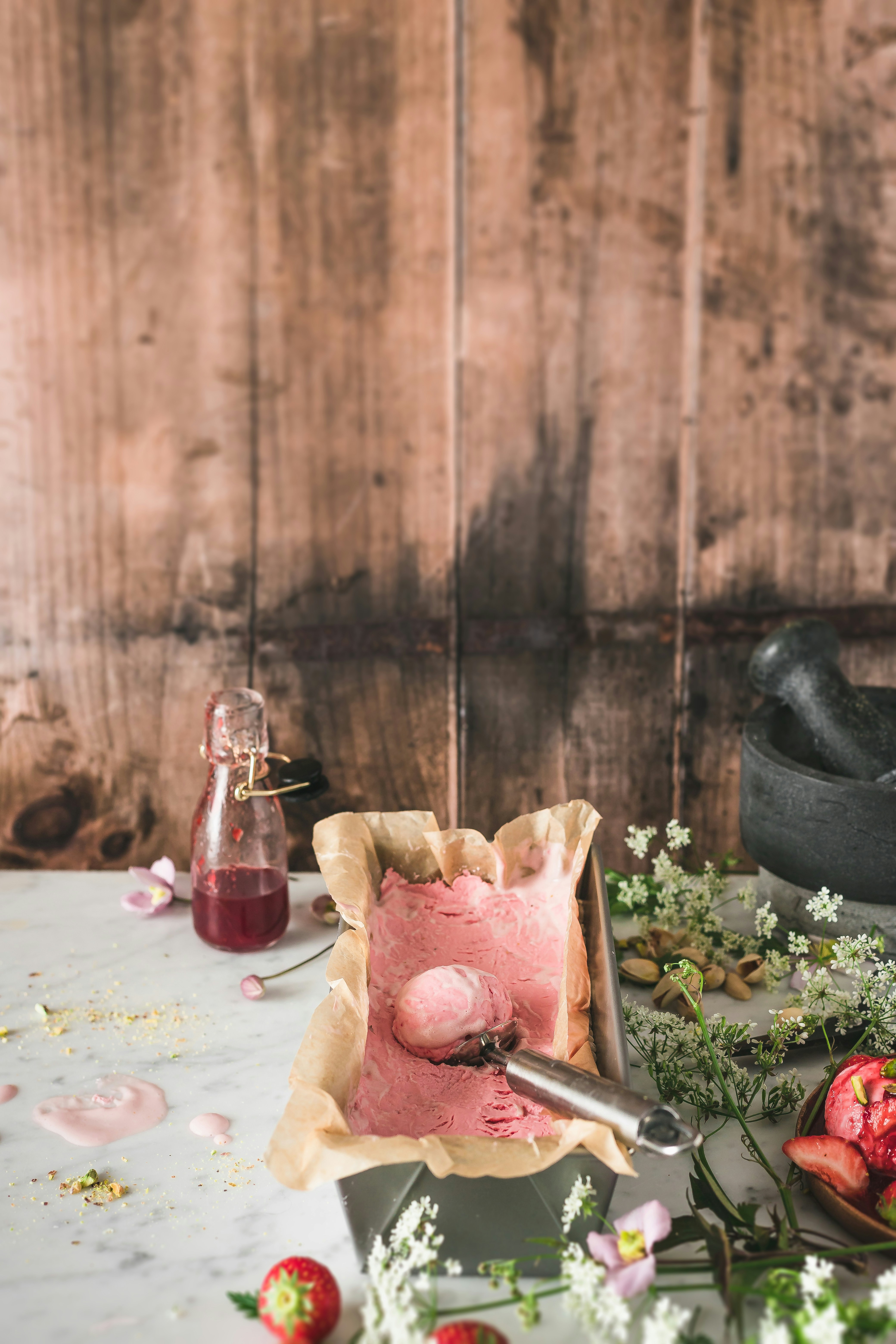 A scoop of strawberry ice cream rests in a parchment-lined container, surrounded by fresh strawberries and delicate flowers on a marble surface.
