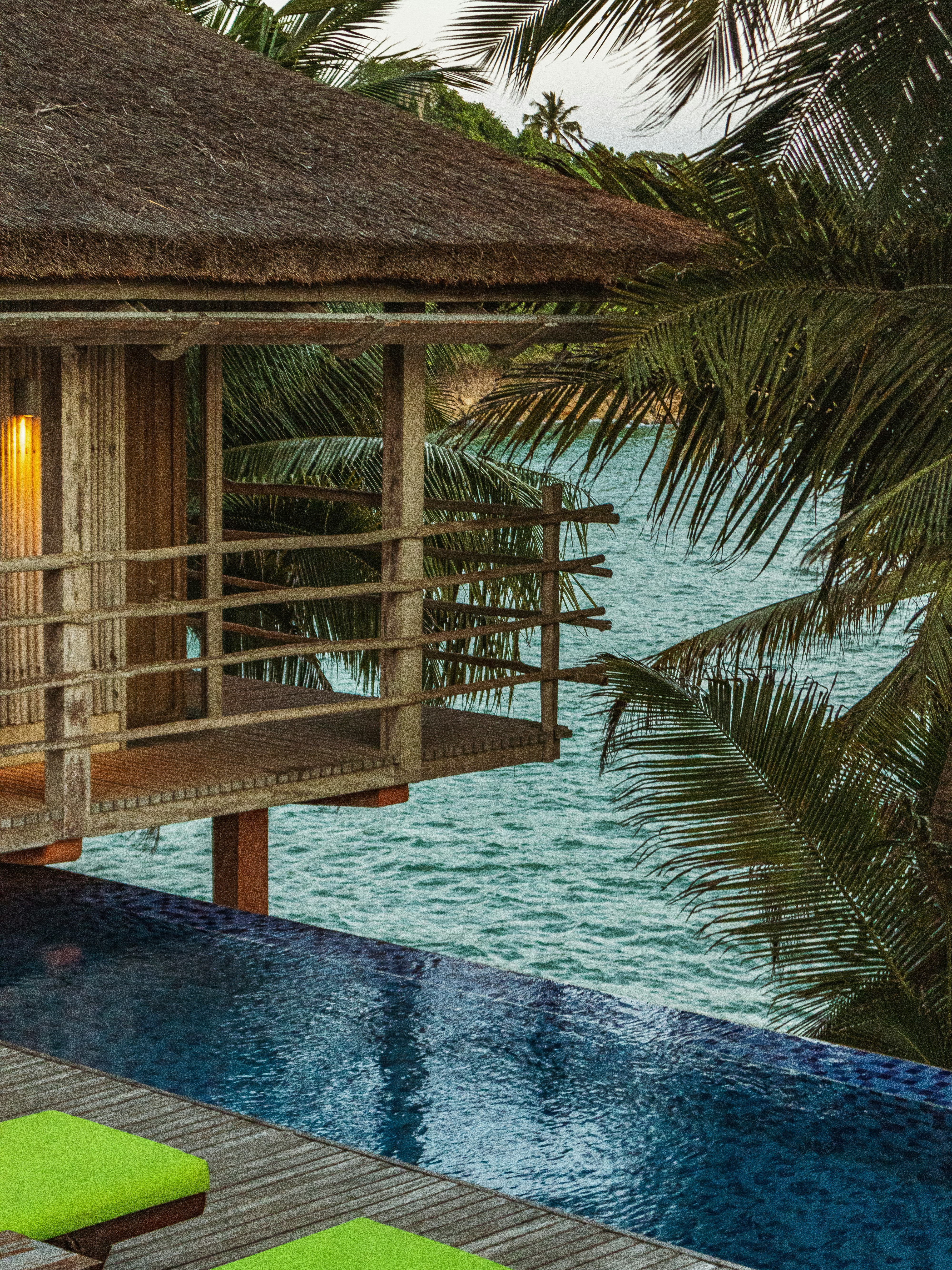 Wooden bungalow overlooking a turquoise sea, framed by lush palm leaves and an infinity pool.