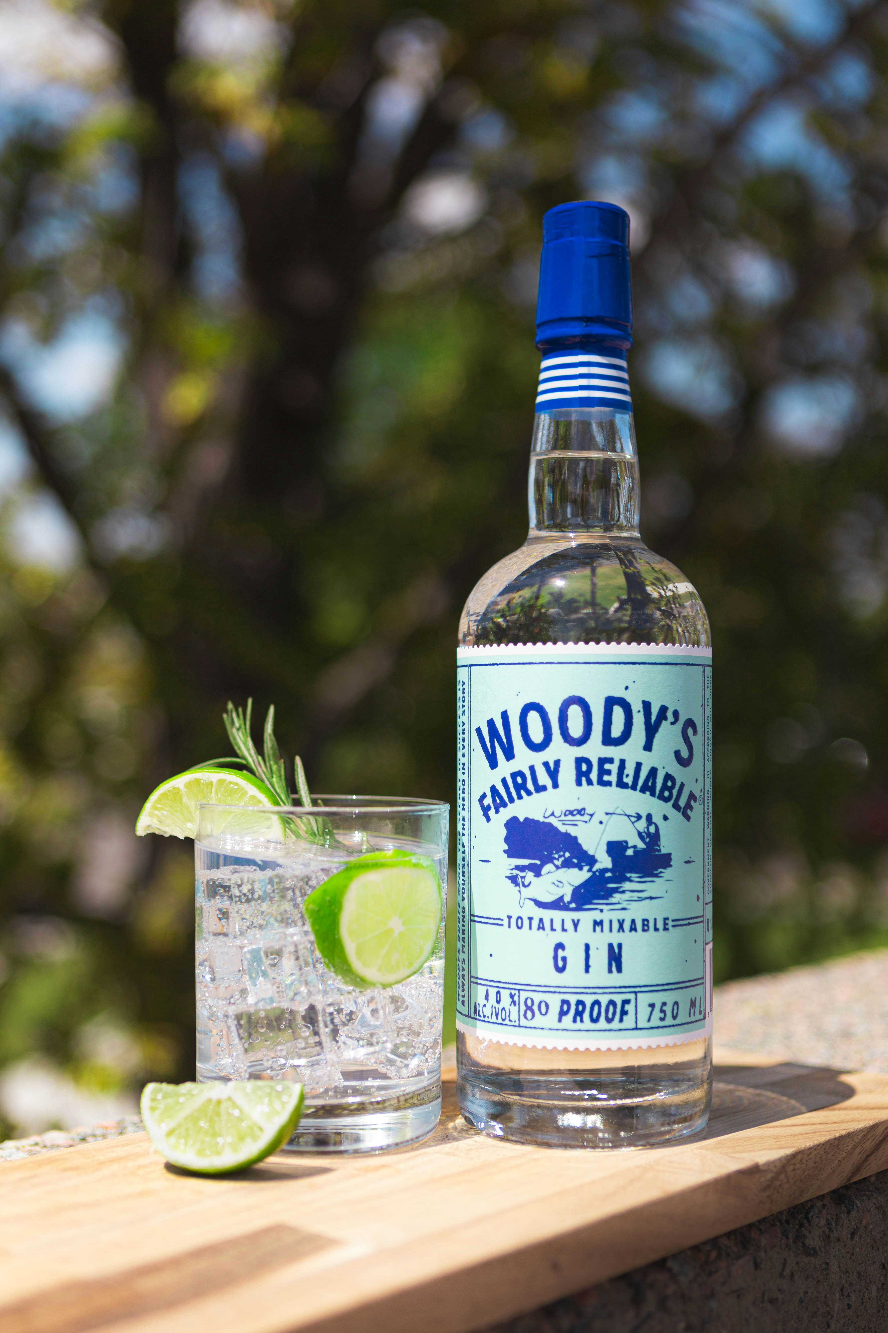 Bottle of Woody's Fairly Reliable gin beside a glass filled with tonic water, garnished with lime and rosemary. The scene captures a sunny outdoor setting.