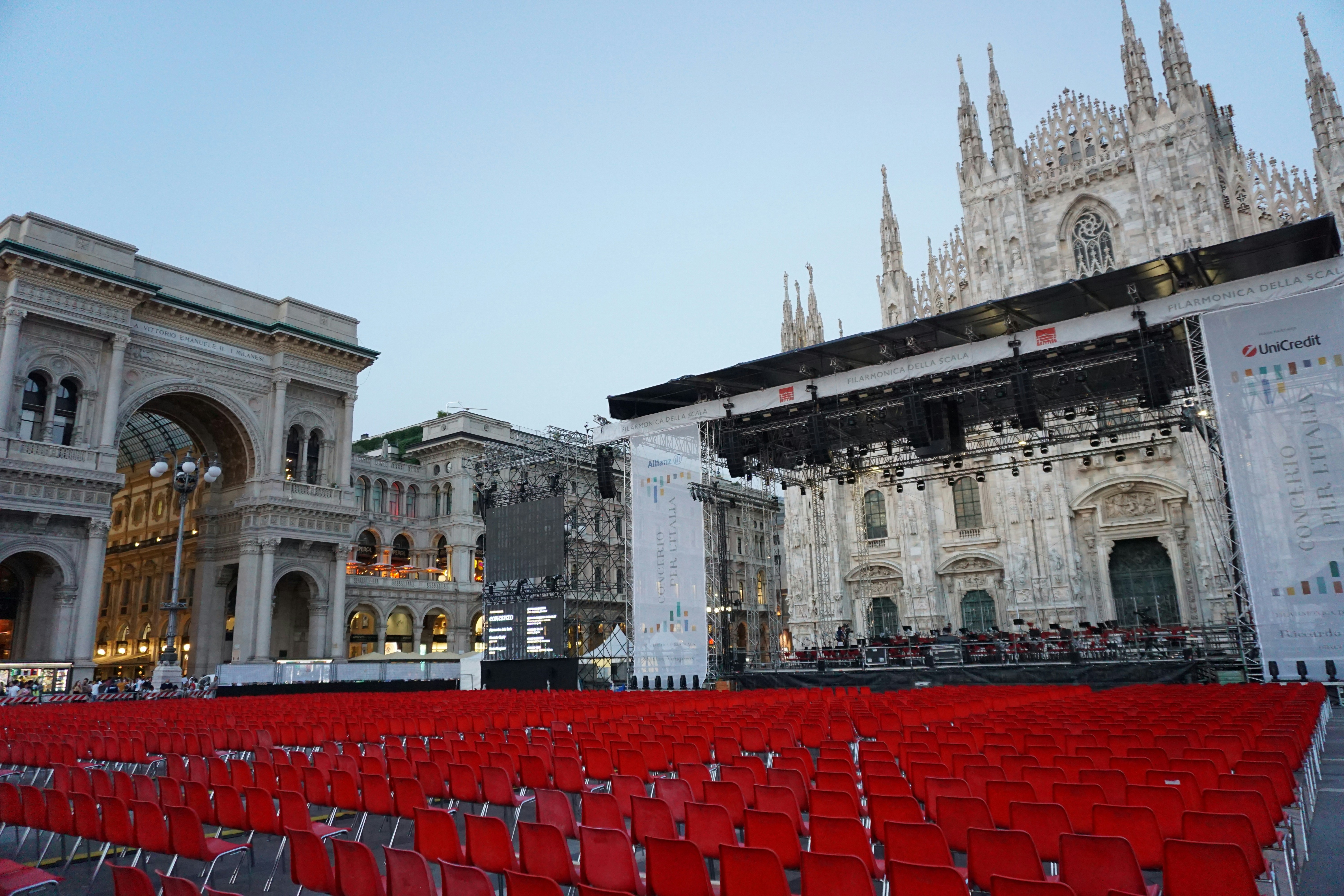 Empty red chairs arranged in front of a grand stage set against the backdrop of a historic cathedral and archway. The scene captures the calm before a lively event.