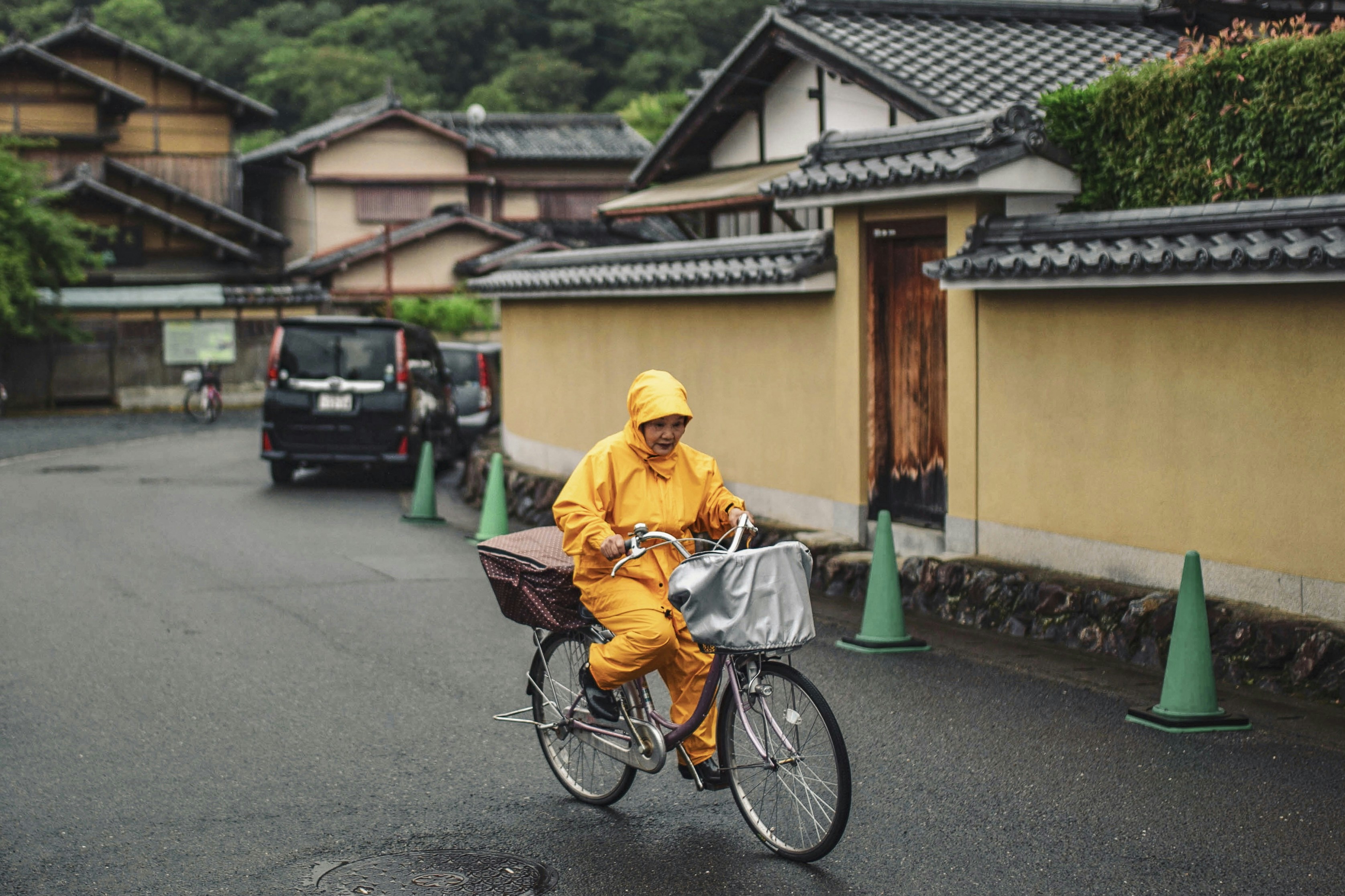 A person in a bright yellow raincoat rides a bicycle along a quiet street, surrounded by traditional Japanese architecture and lush greenery.