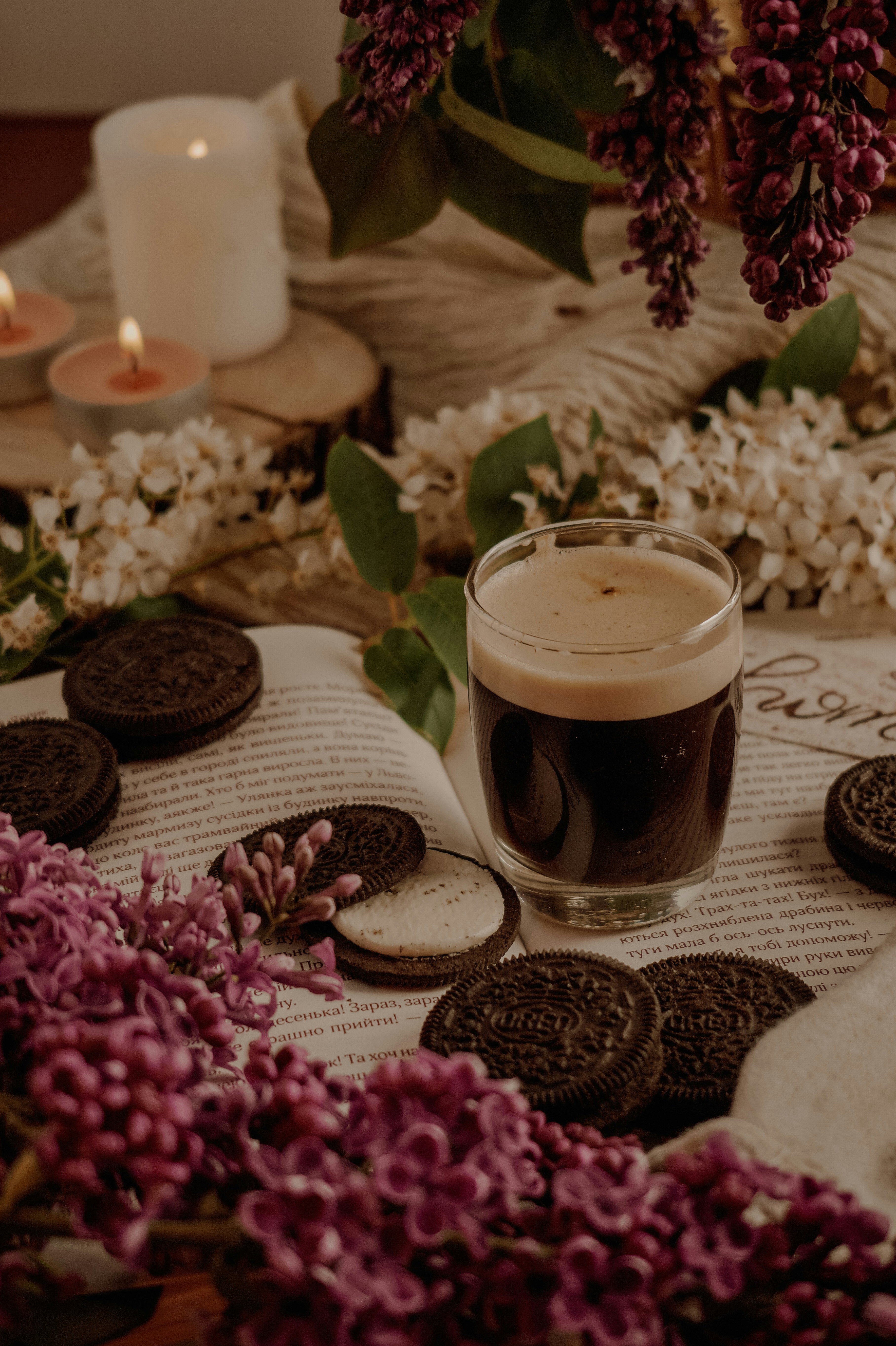 A glass of dark espresso surrounded by Oreo cookies and vibrant lilac flowers, resting on an open book. Soft candlelight adds warmth to the scene.