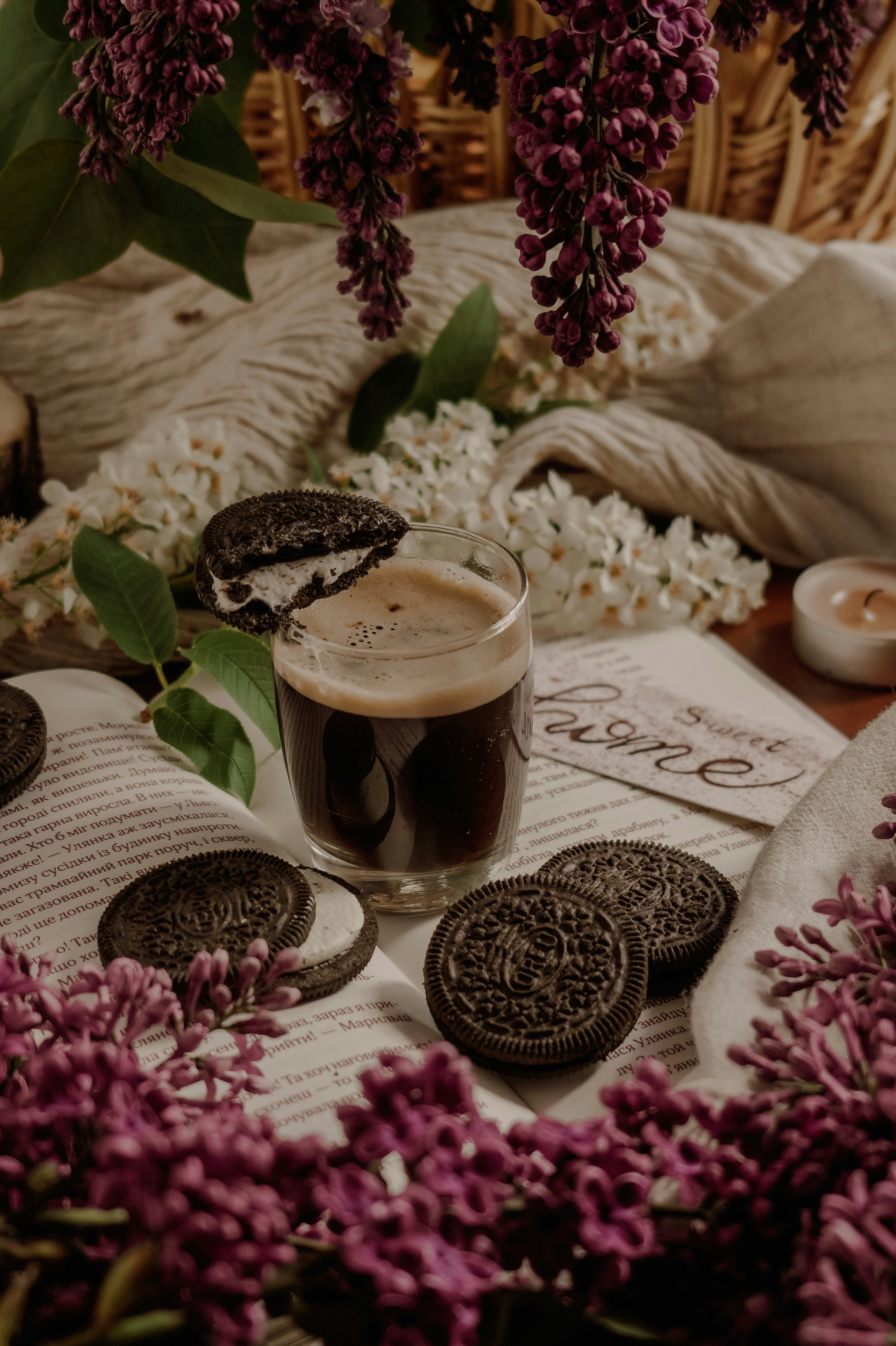 A glass of dark espresso sits beside Oreo cookies on a folded newspaper, framed by hanging lilacs and soft fabric for a cozy setup.
