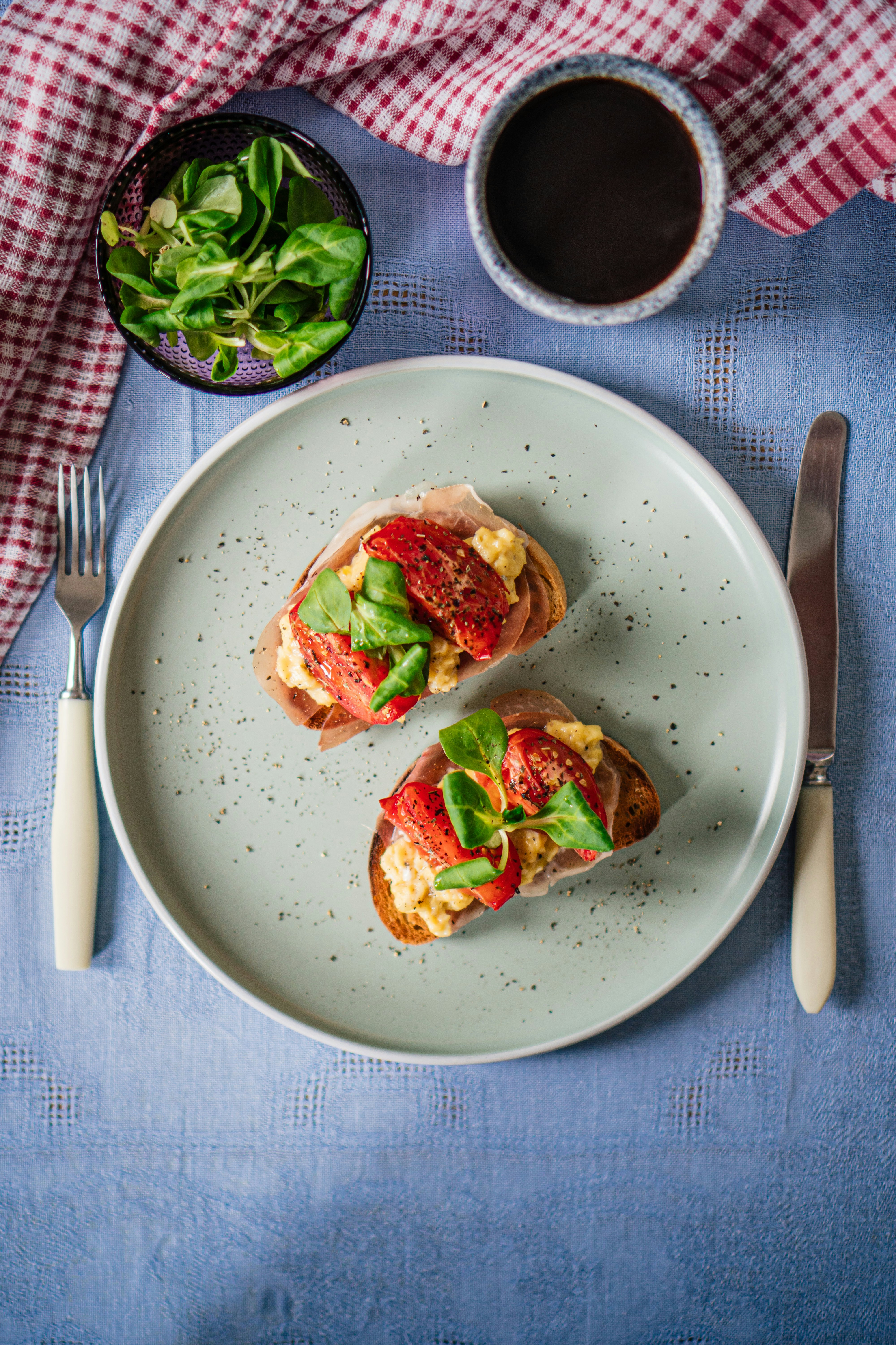 Artfully arranged toast topped with scrambled eggs, roasted tomatoes, and fresh basil, accompanied by a side of greens and coffee.