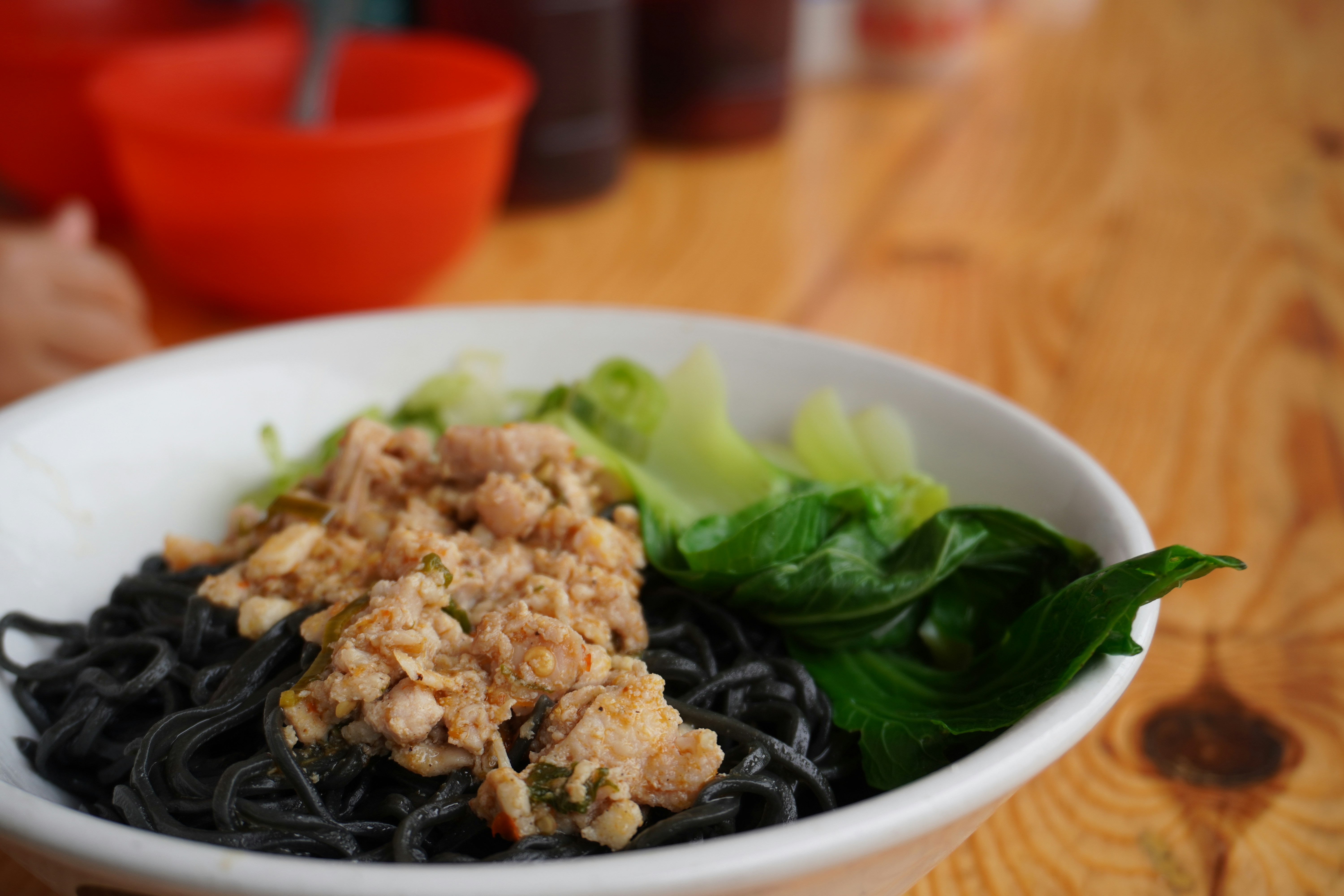 Bowl of black noodles topped with minced chicken and fresh greens on a wooden table.