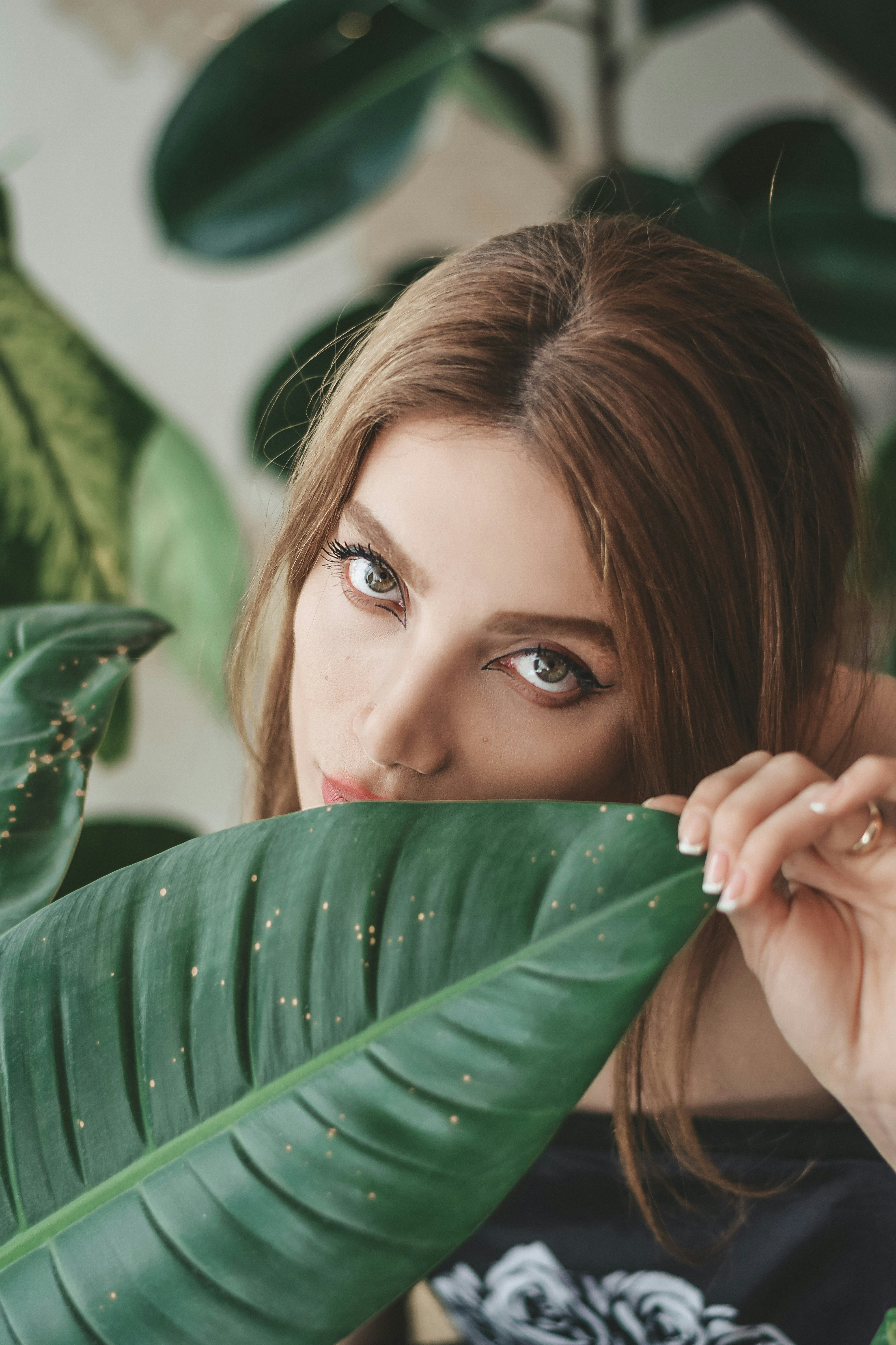 A young woman partially obscured by a large green leaf, gazing directly at the viewer amidst lush foliage. Her expressive eyes and gentle pose create a connection with nature.