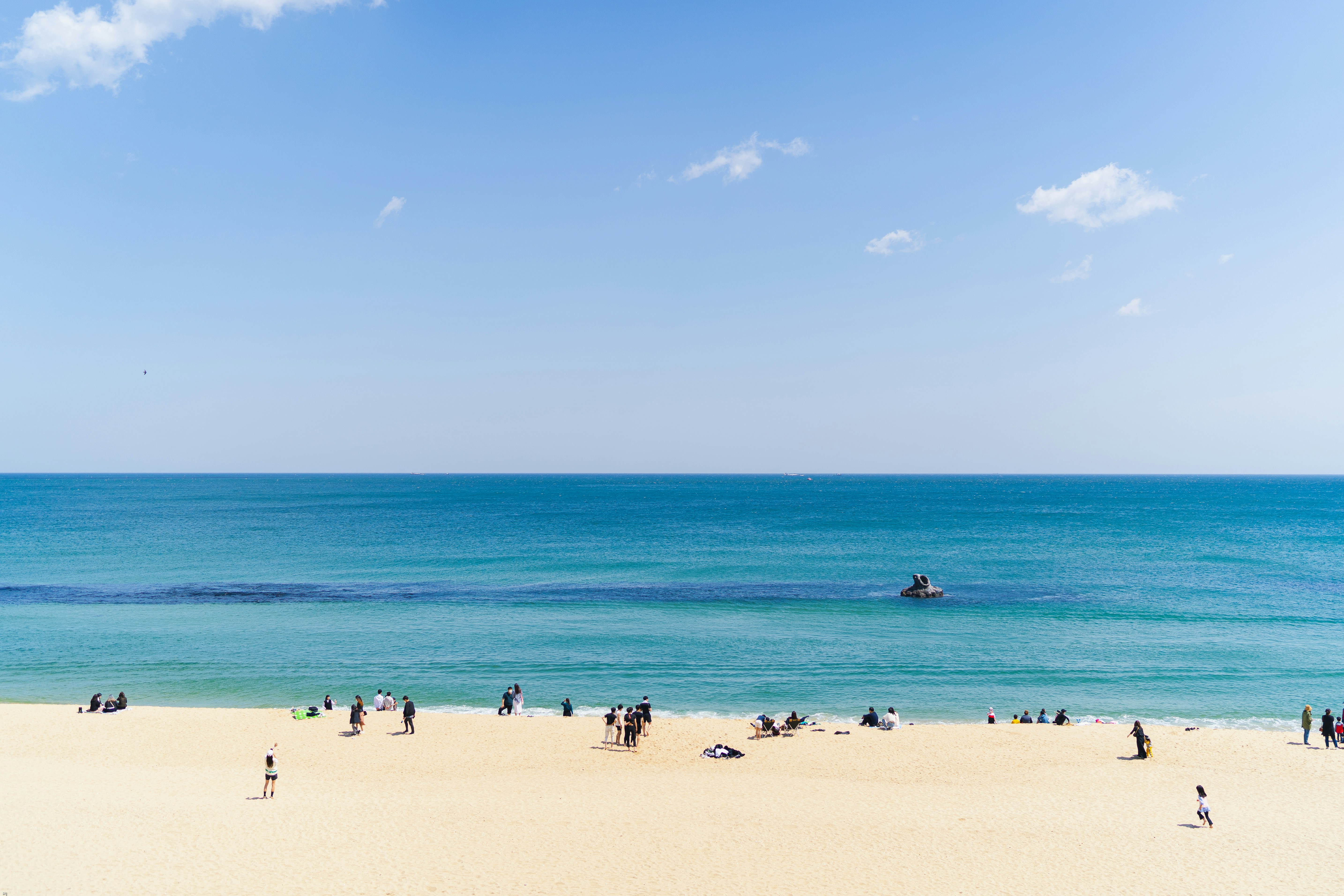 Crowd enjoying a sunny day on the beach with gentle waves lapping at the shore. A small boat is visible in the distance.