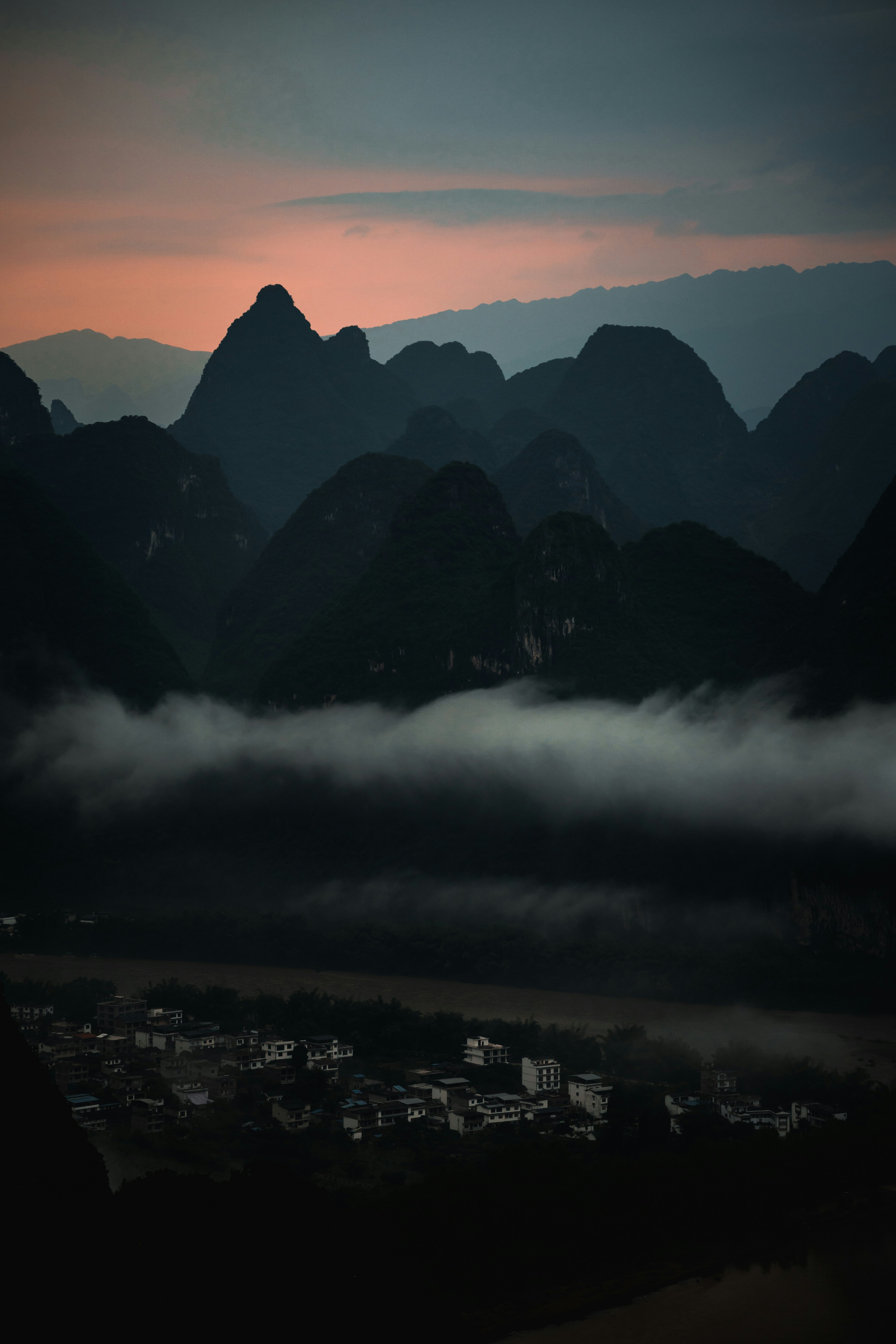 Misty mountains loom over a tranquil village, enveloped in a soft twilight glow. The scene captures the serene beauty of nature's silhouettes.