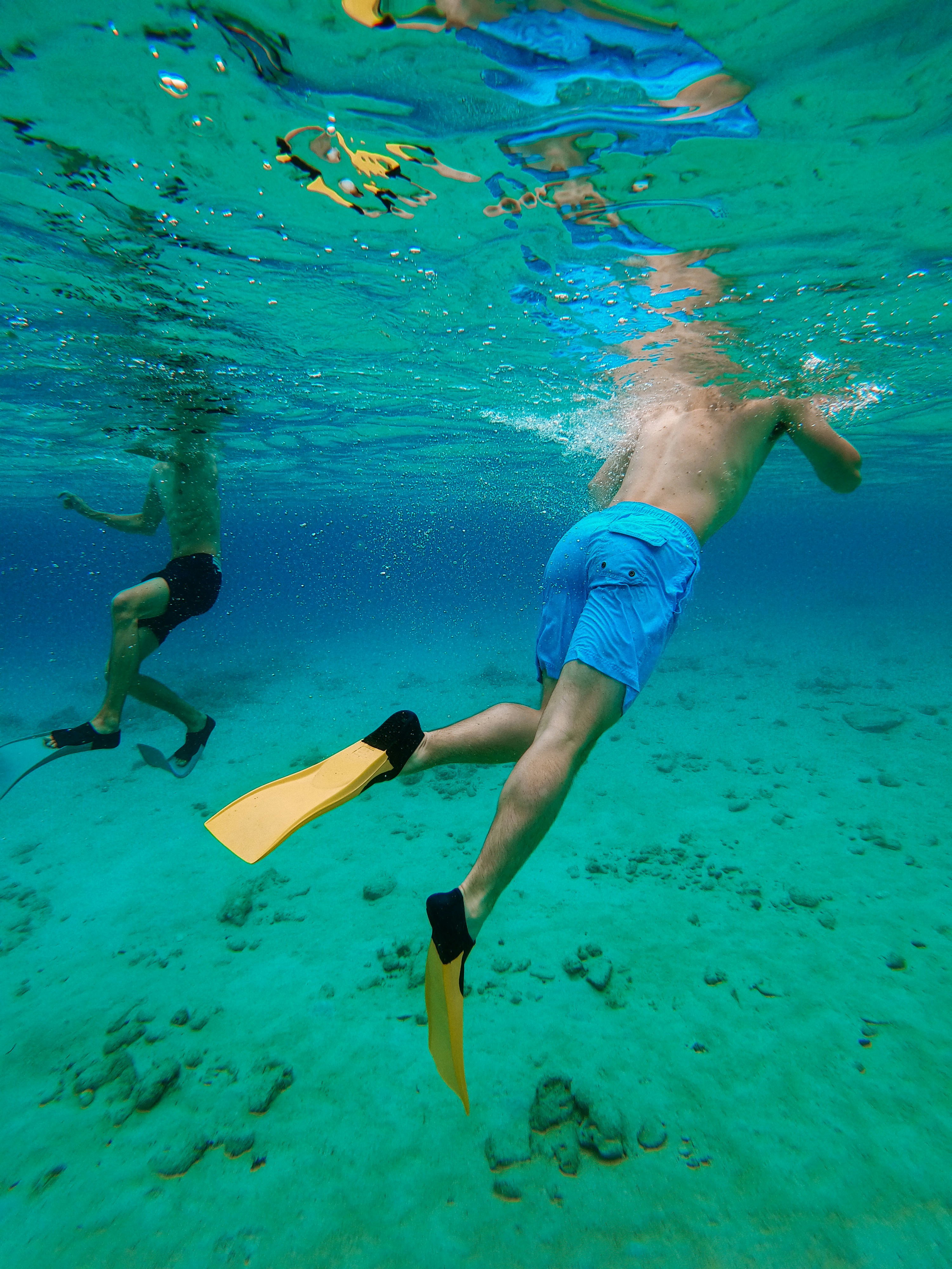 Underwater photograph of two divers gliding over a sandy seabed; foreground diver wears blue swim shorts and yellow fins as bubbles rise.