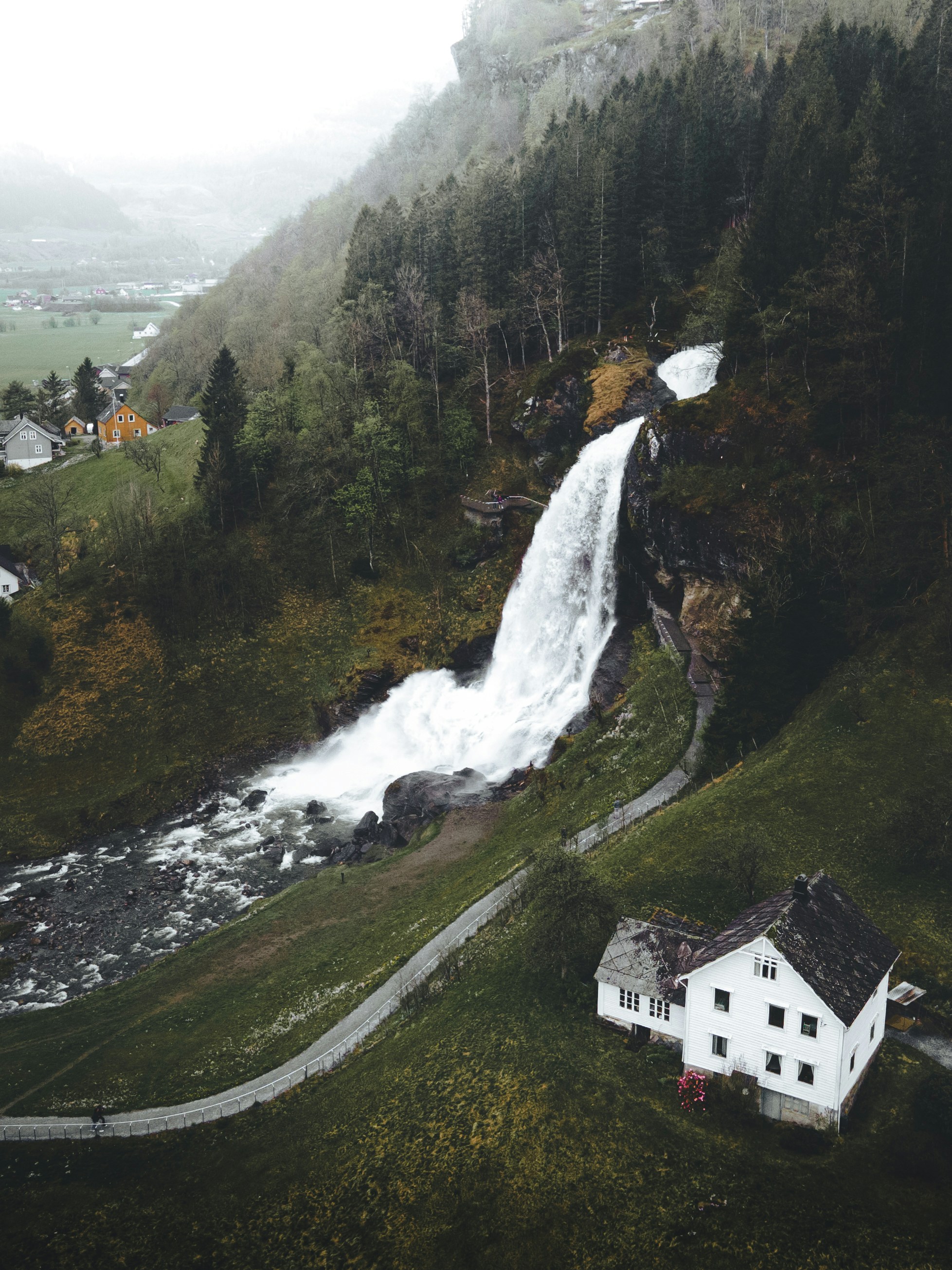A majestic waterfall flows down a rocky cliff, surrounded by lush greenery and a quaint house nearby. The scene captures the serene beauty of nature's power.