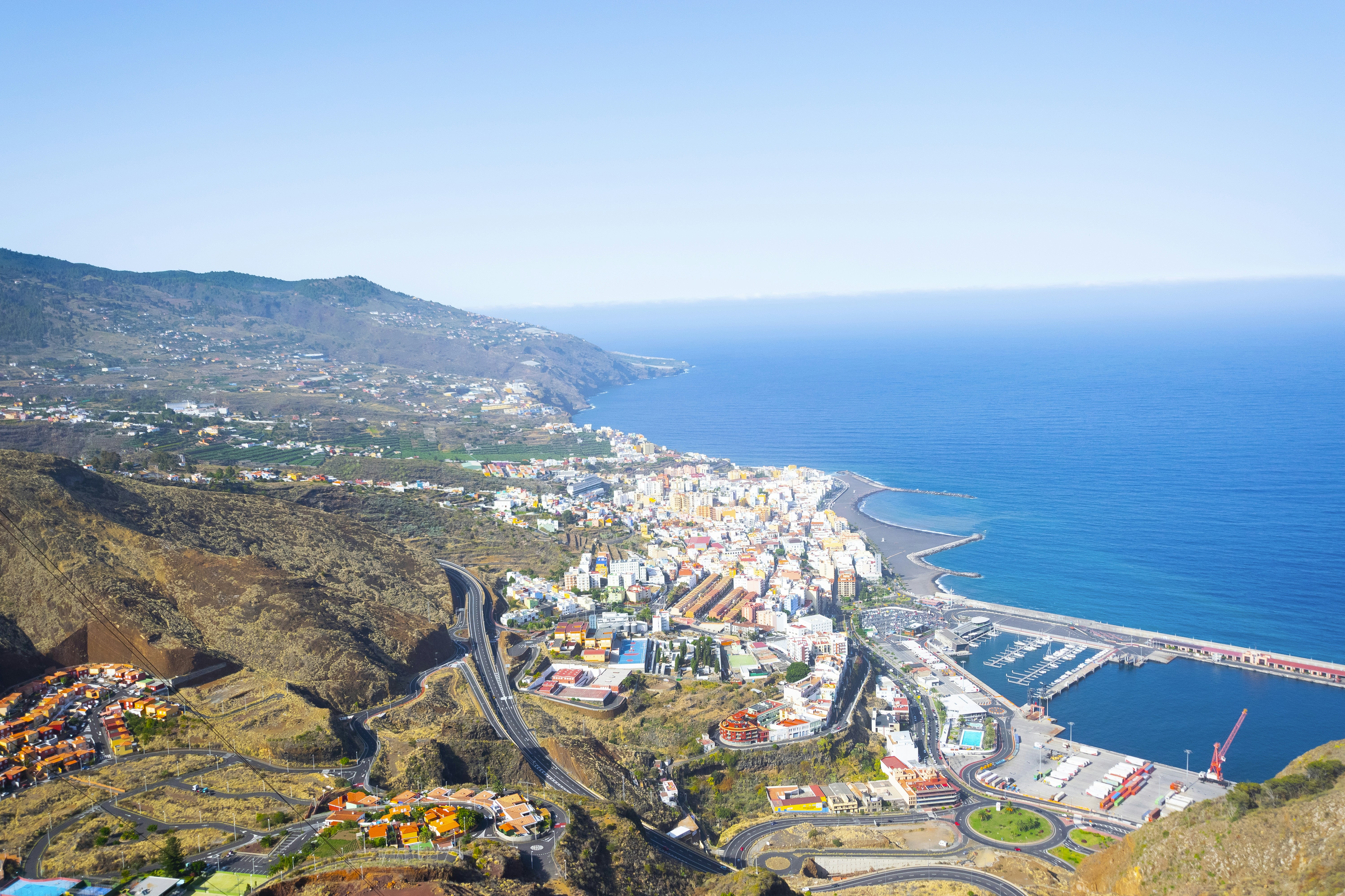 Aerial view of a coastal city nestled between rugged mountains and the expansive blue ocean under a clear sky.