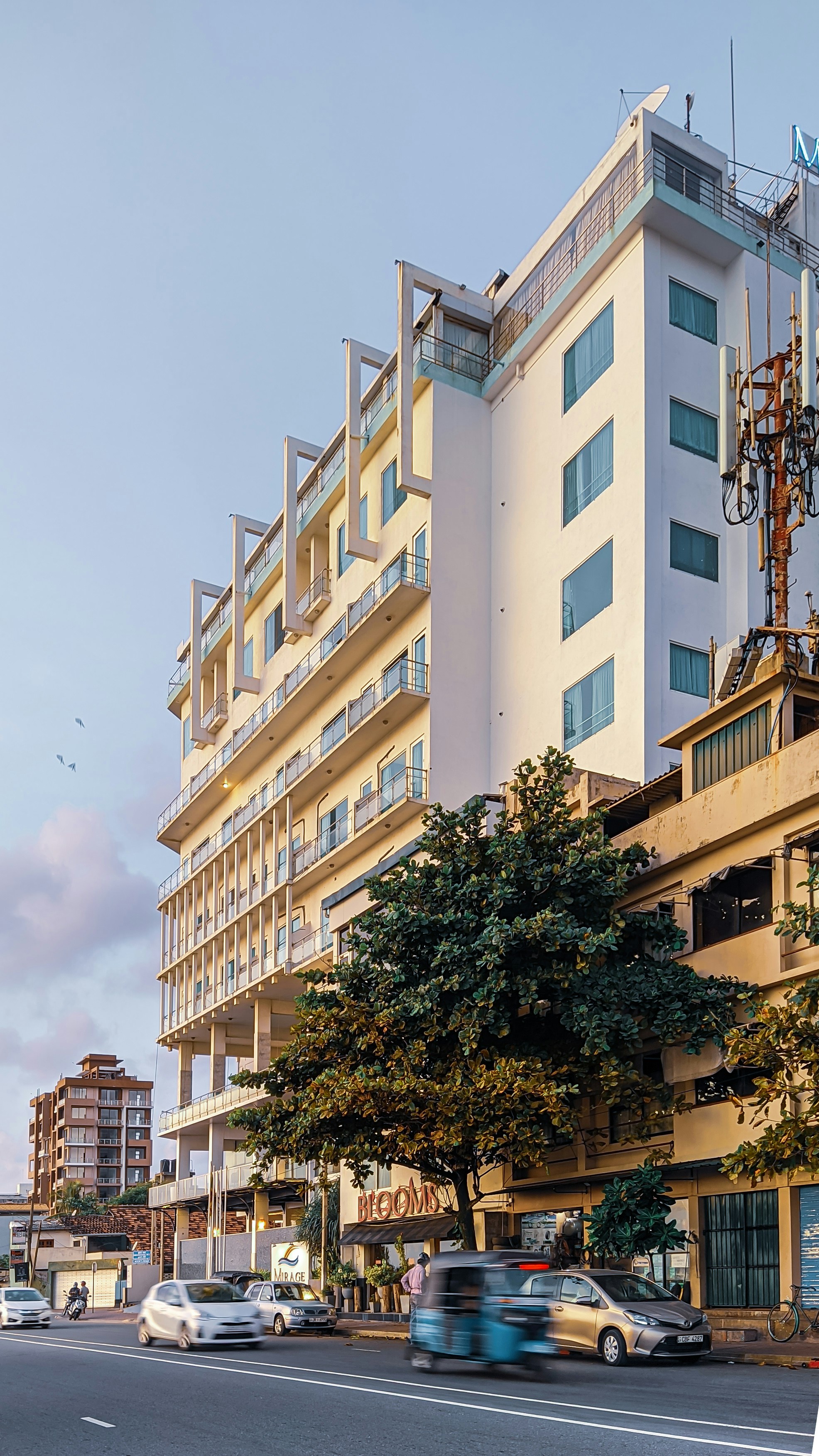 Tall modern building with angular balconies against a clear sky, bordered by a busy street with motion-blurred vehicles.