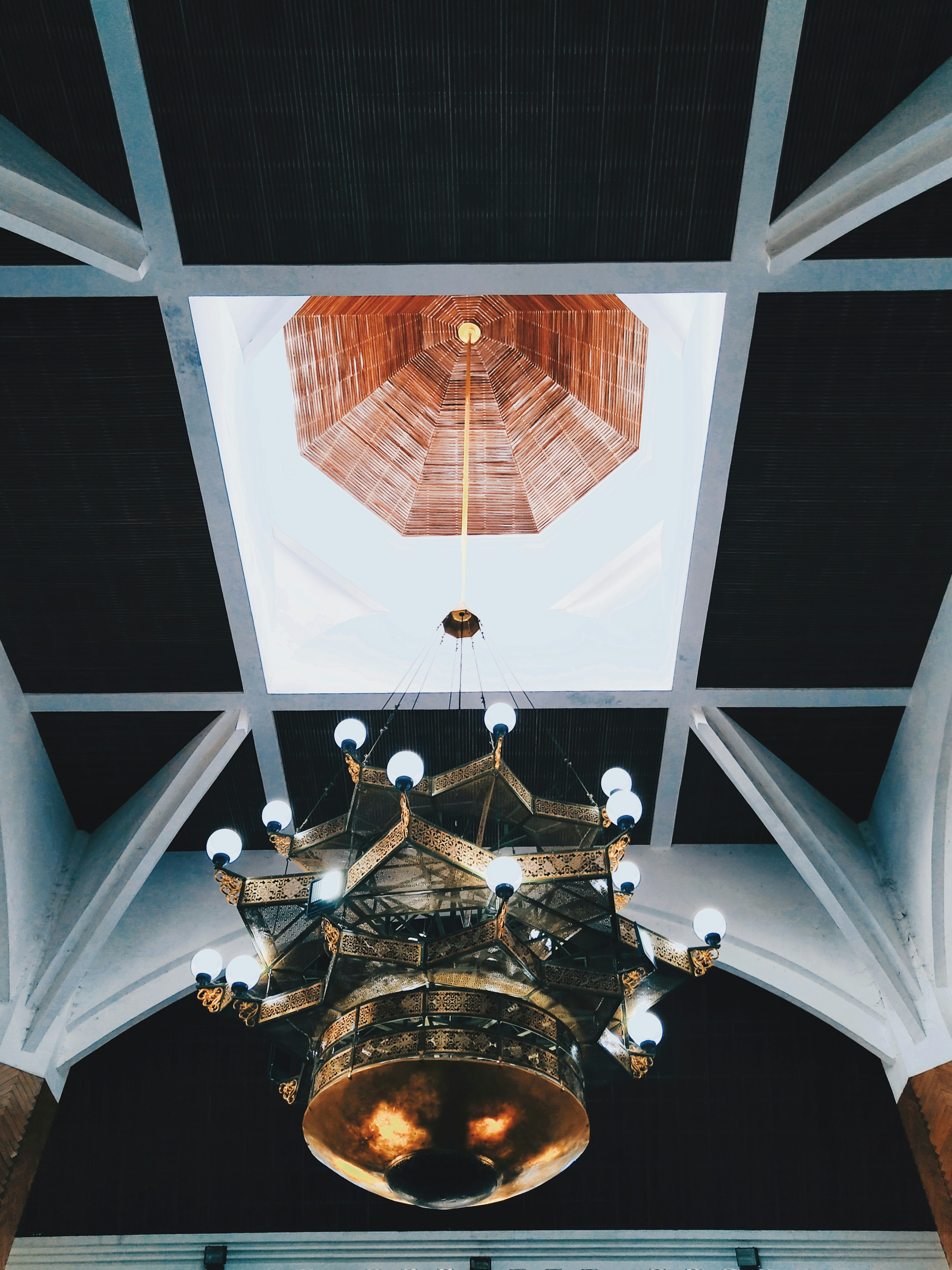 Intricate chandelier hanging from a geometric ceiling, framed by a skylight that reveals a wooden structure above.