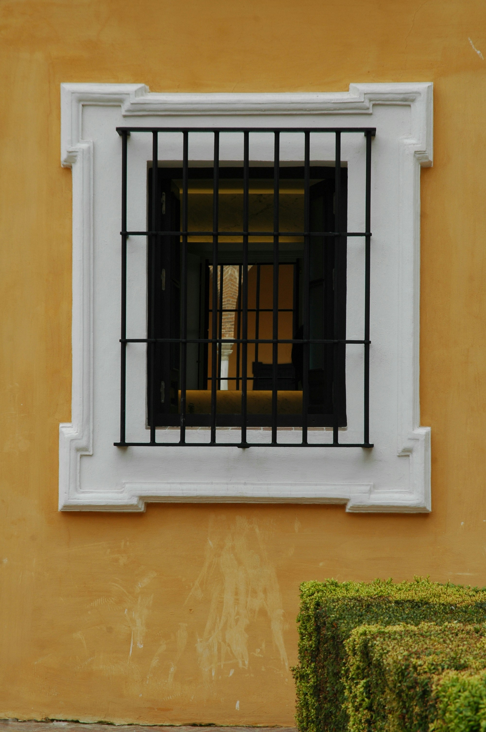 A black barred window set against a vibrant yellow wall, revealing a softly lit interior space. The surrounding greenery adds a touch of nature's calm.