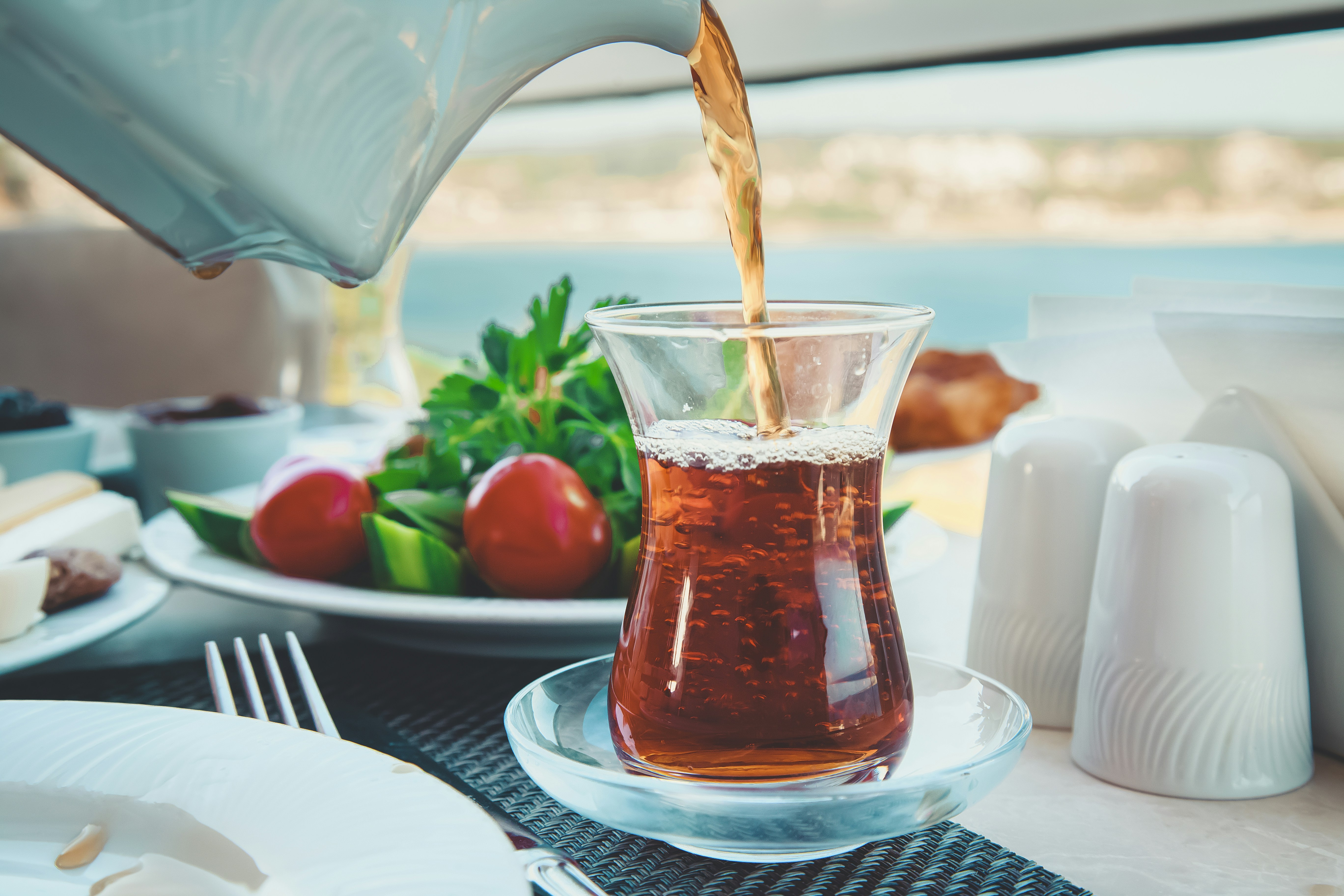 Tea being poured into a delicate glass alongside a fresh salad and assorted dishes, with a picturesque background of water and hills.