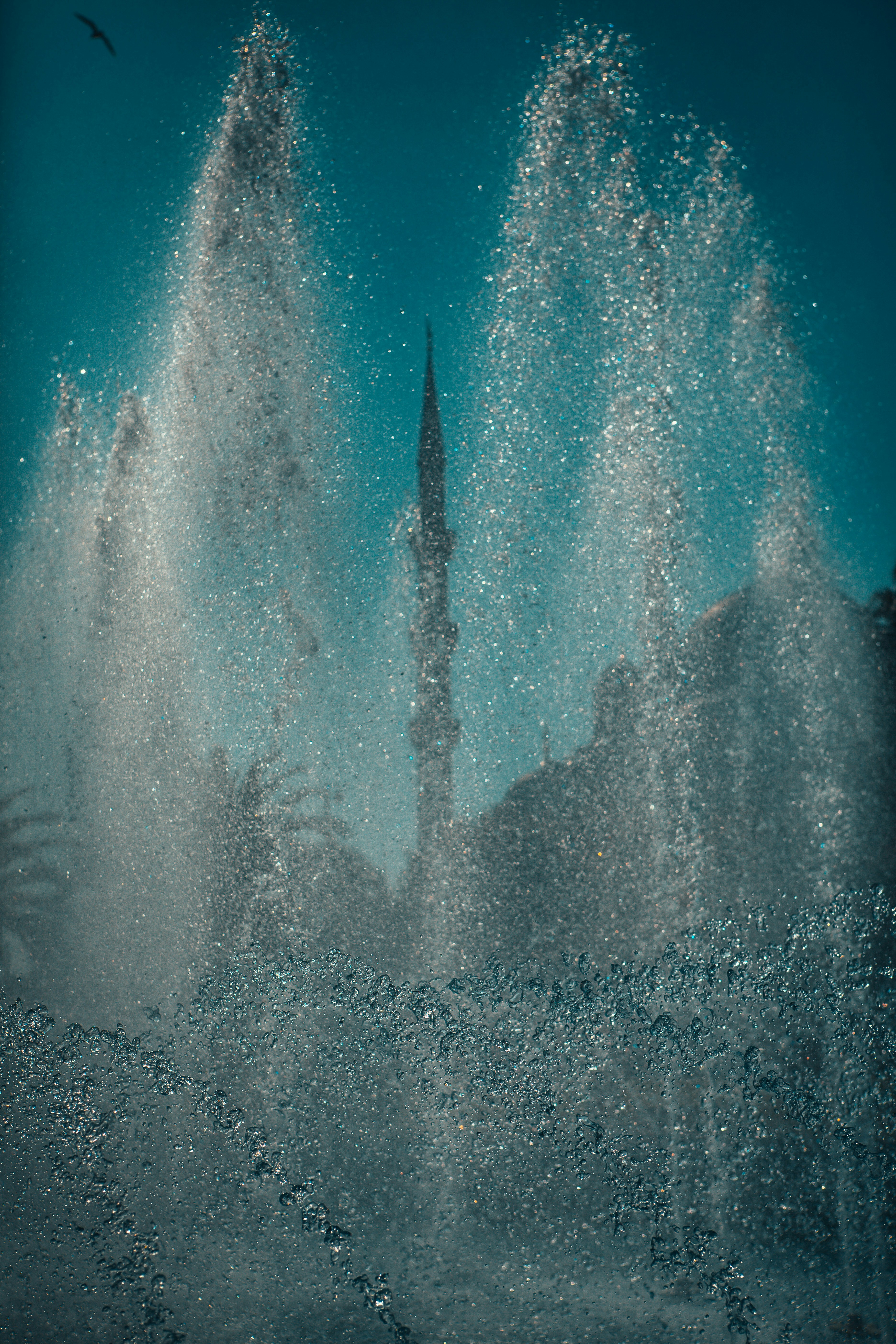 Water fountains erupting in a vibrant display against a clear blue sky, with a silhouette of a spire in the background.