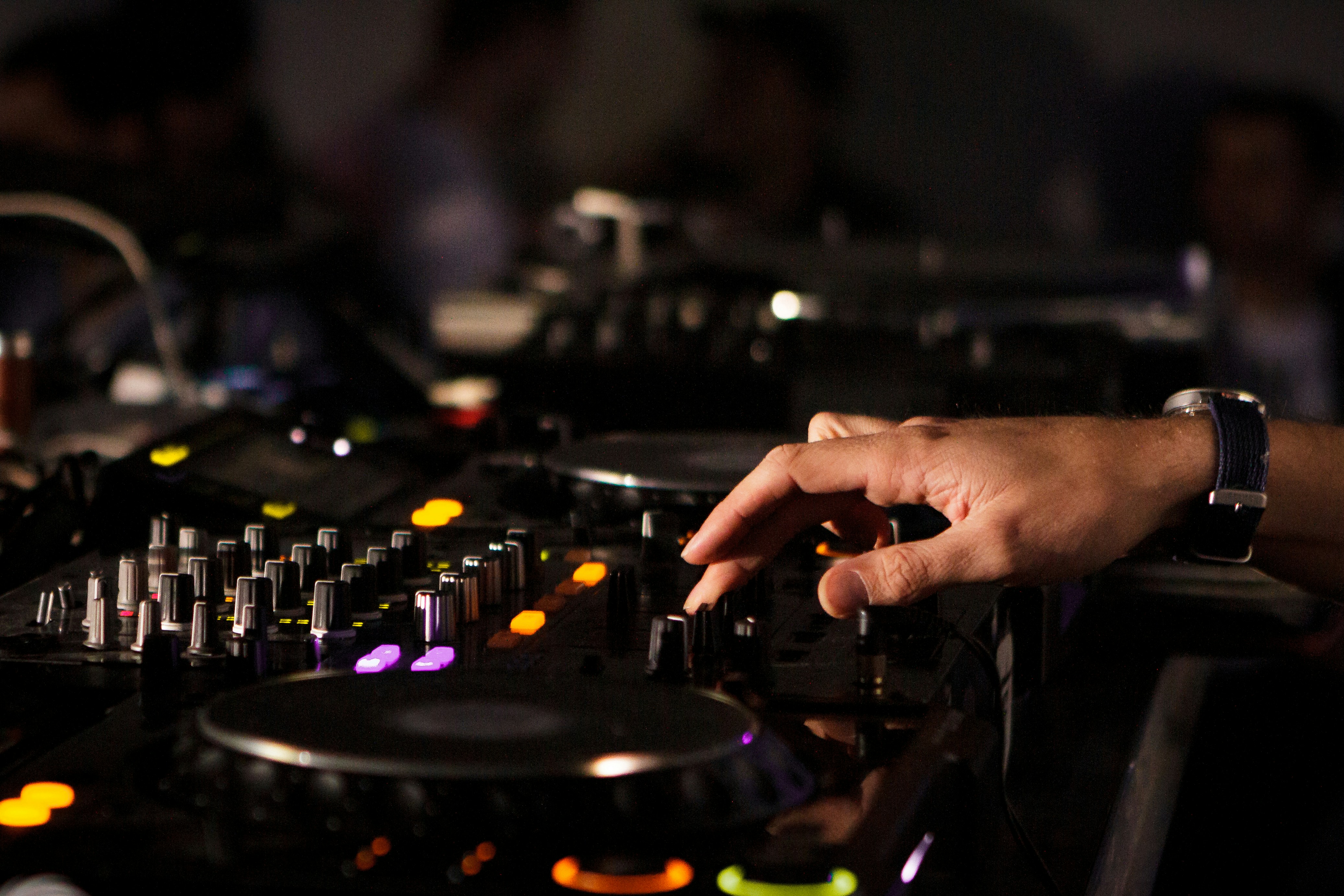 A DJ's hand skillfully adjusts the controls on a mixing console, surrounded by a vibrant atmosphere of a live music event.