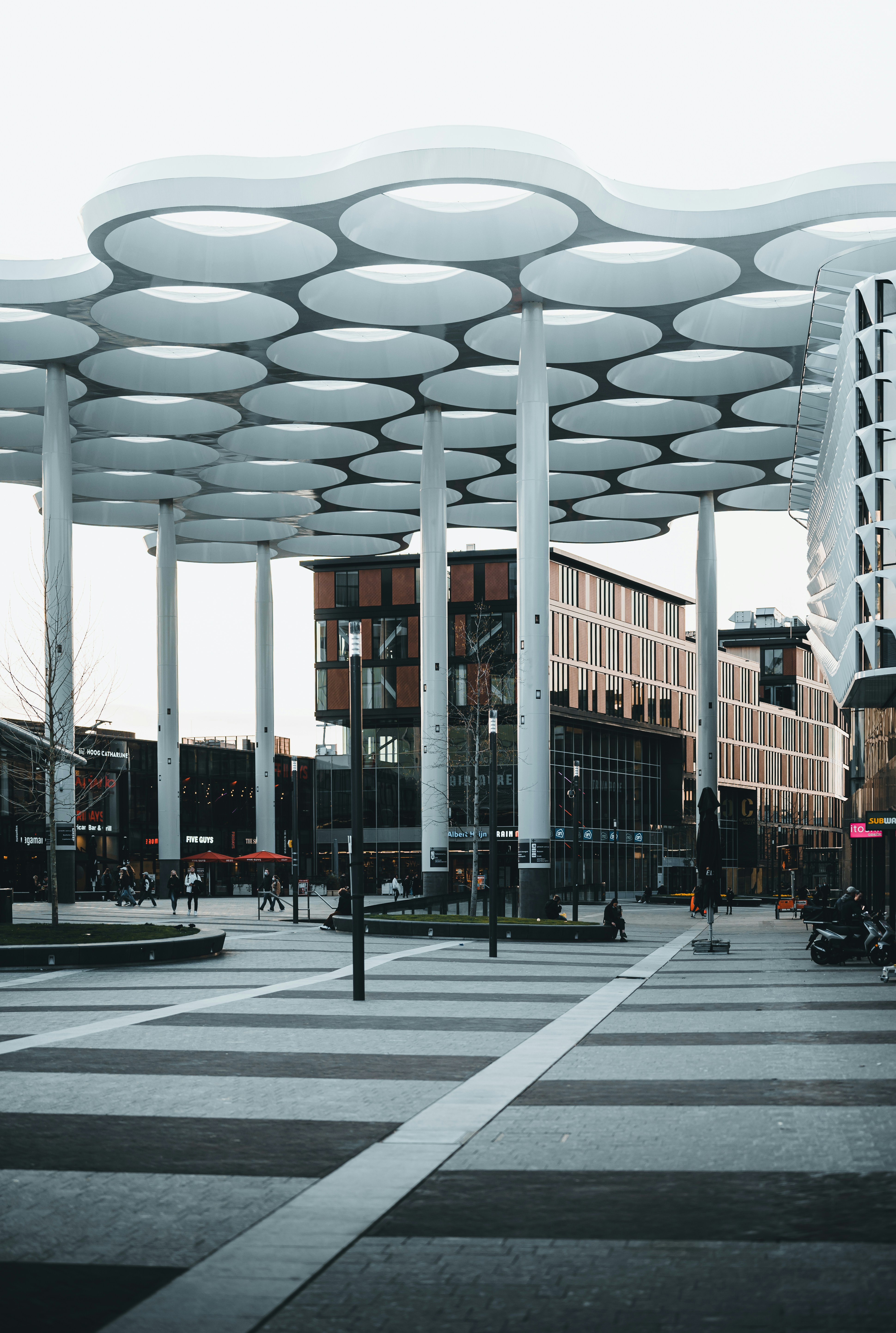 Architectural design featuring a unique, cloud-like canopy supported by tall white columns, framing a bustling urban square with contemporary buildings.
