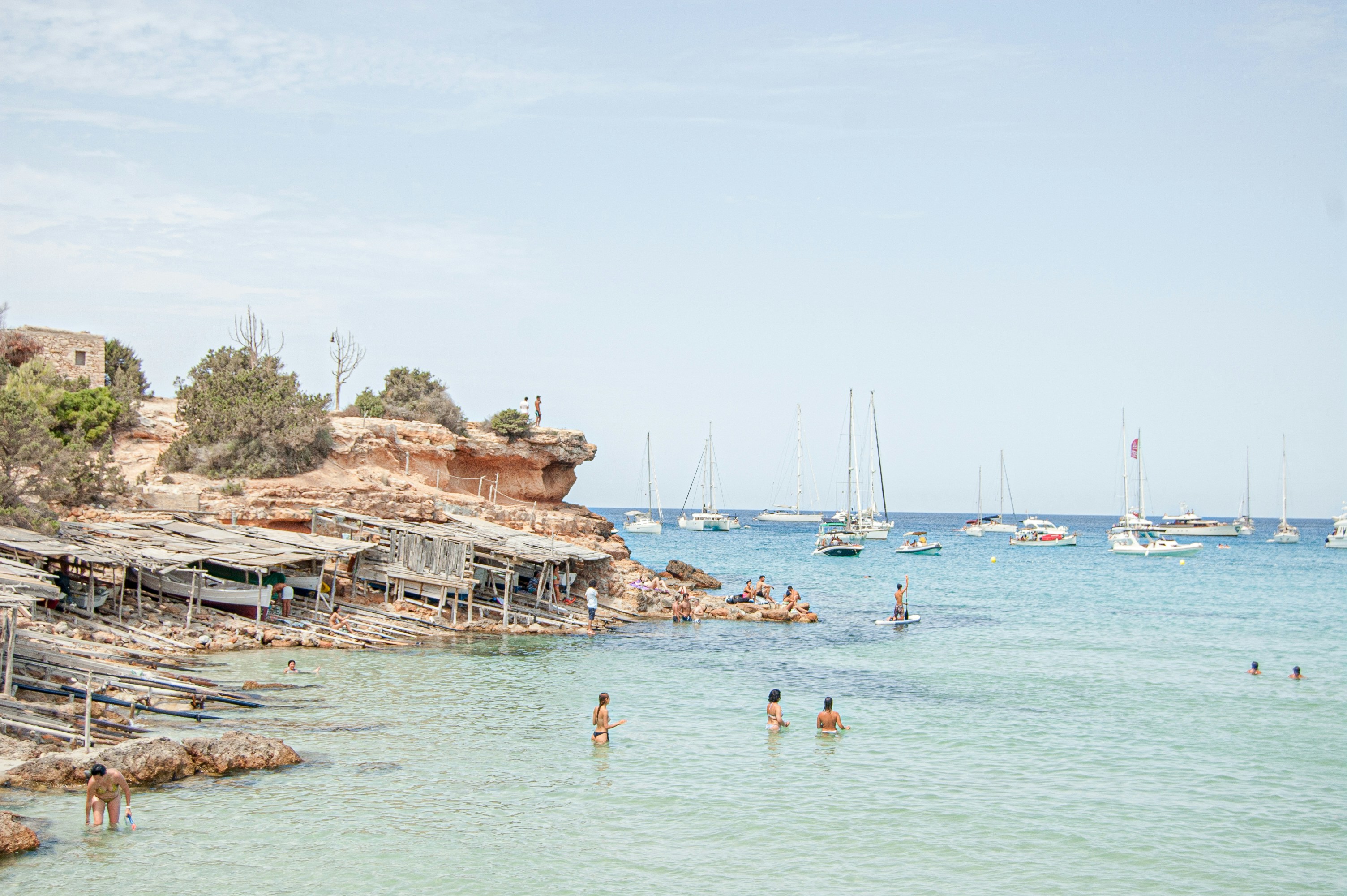 Calm beach scene featuring swimmers and paddleboarders near a rocky shoreline with boats in the background.