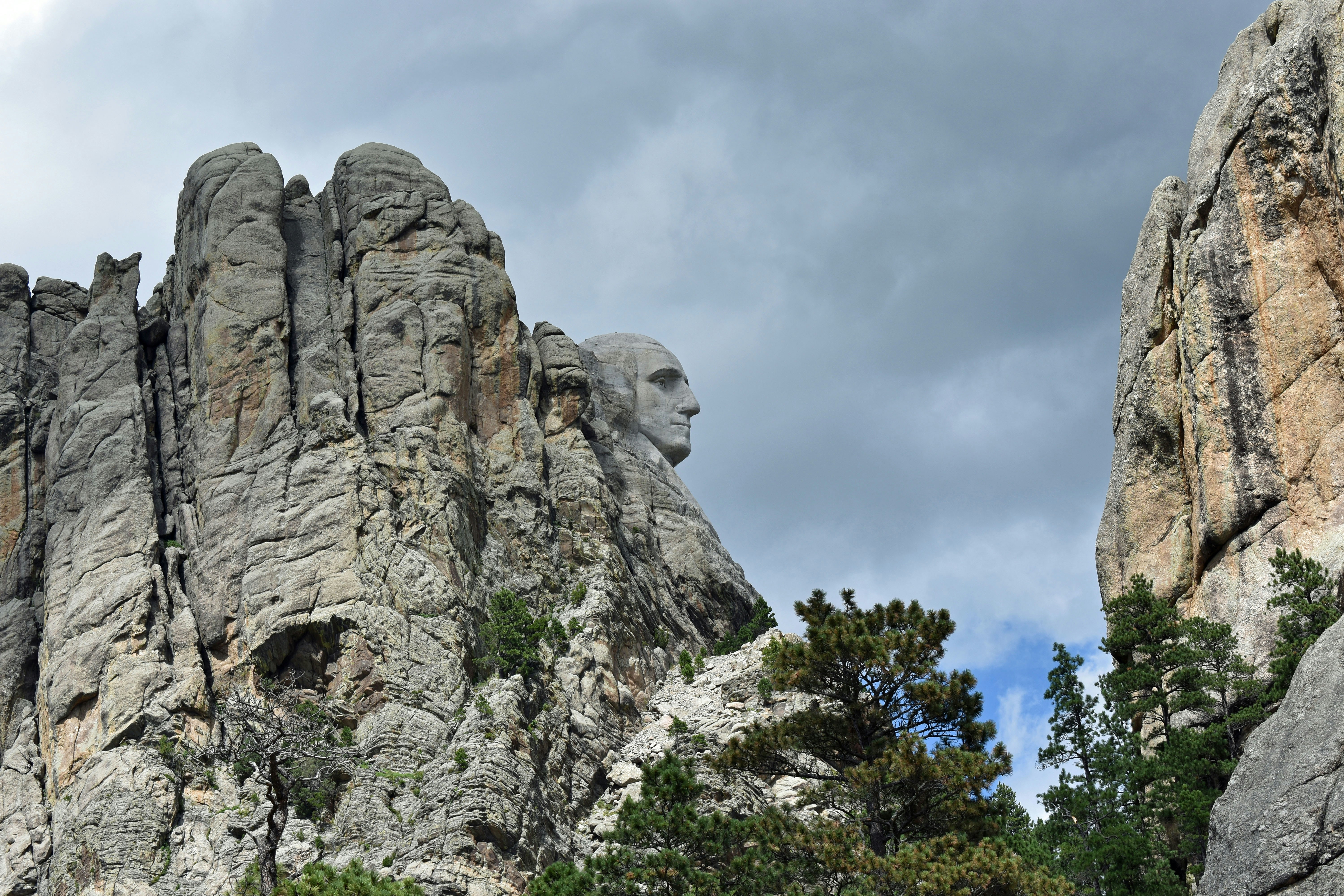 Carved presidential visage emerging from rugged cliffs, surrounded by pine trees under a moody sky.