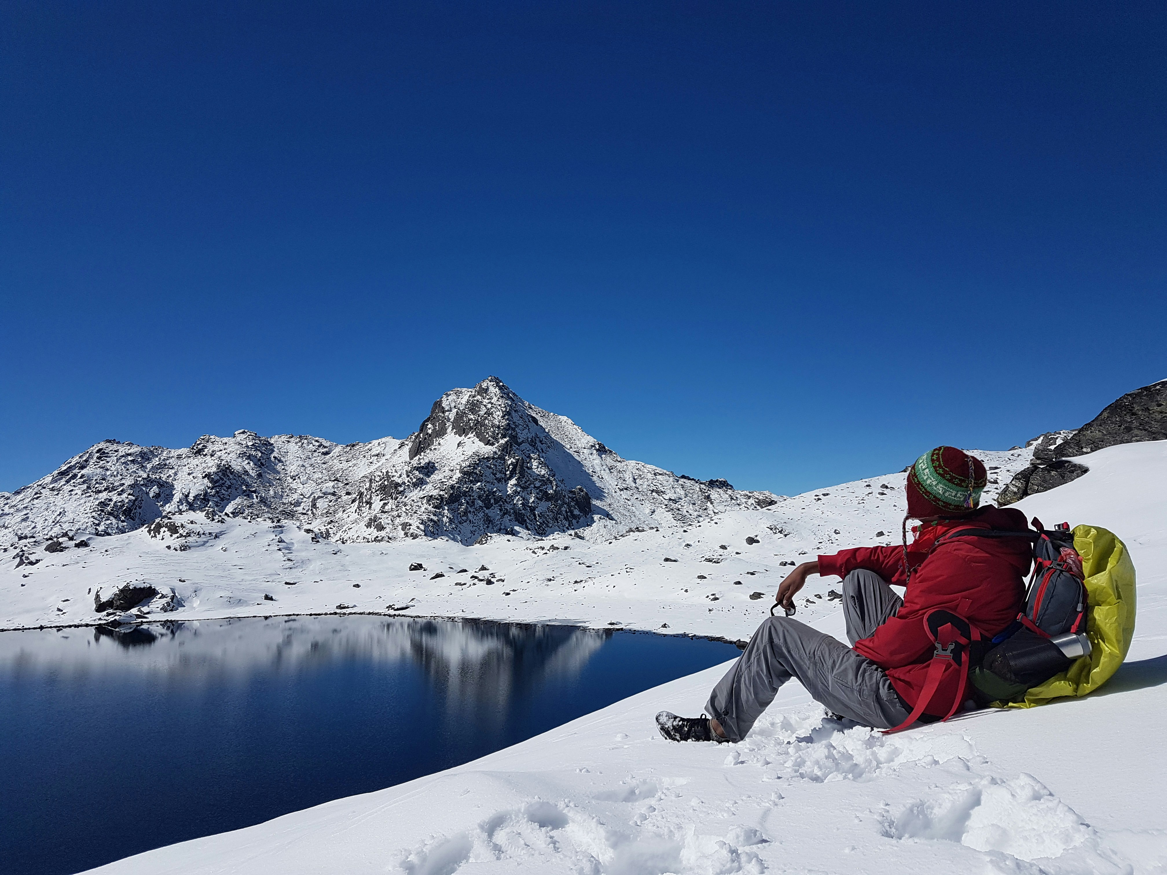A hiker in a red jacket sits on a snowy slope, gazing at a serene mountain lake surrounded by rugged peaks under a clear blue sky.