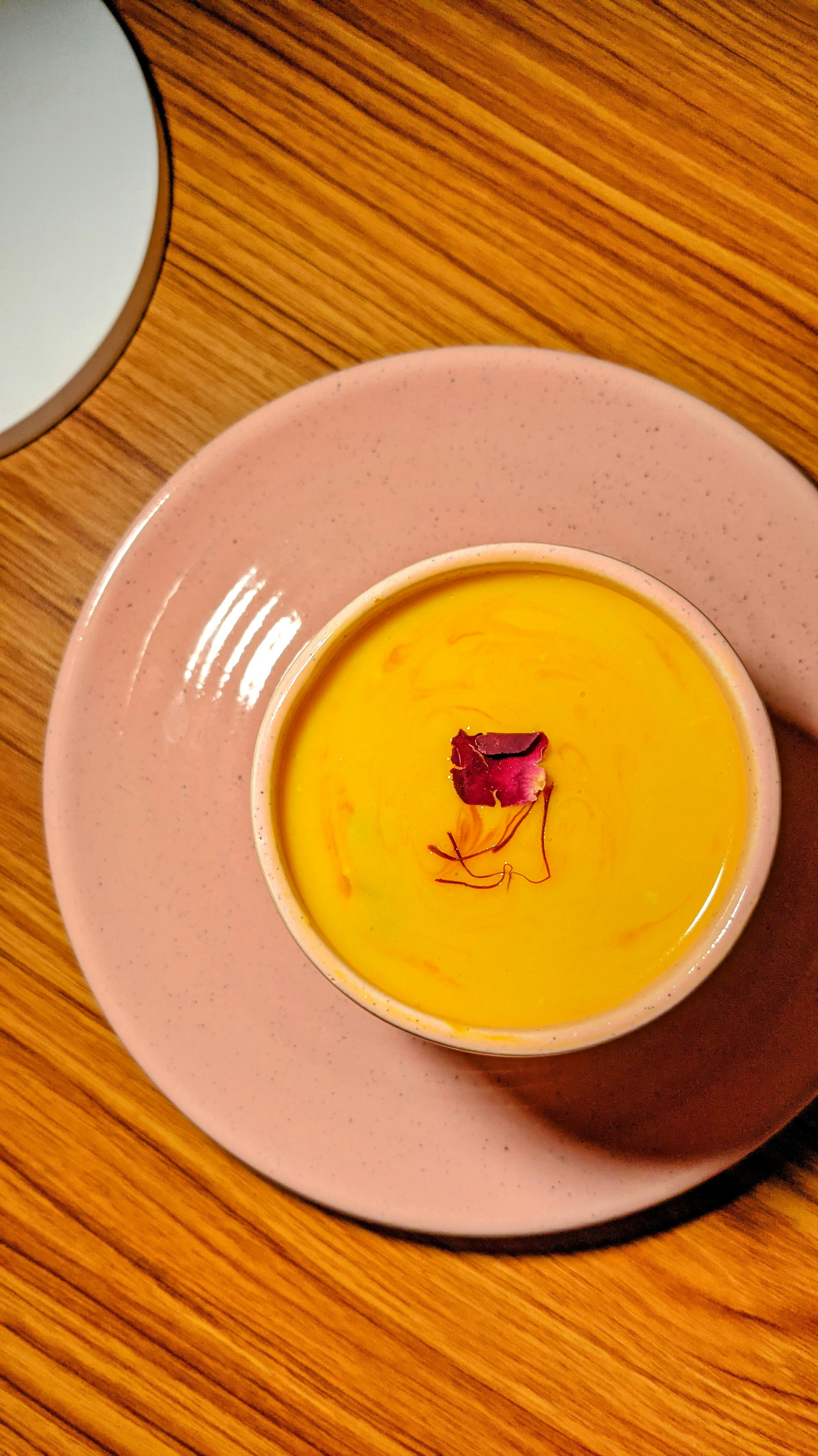 Pink ceramic plate holds a small bowl of yellow cream with a lone red petal floating on its surface, resting on a warm wooden table. The close-up emphasizes the creamy texture and vivid color contrast against the pink plate.