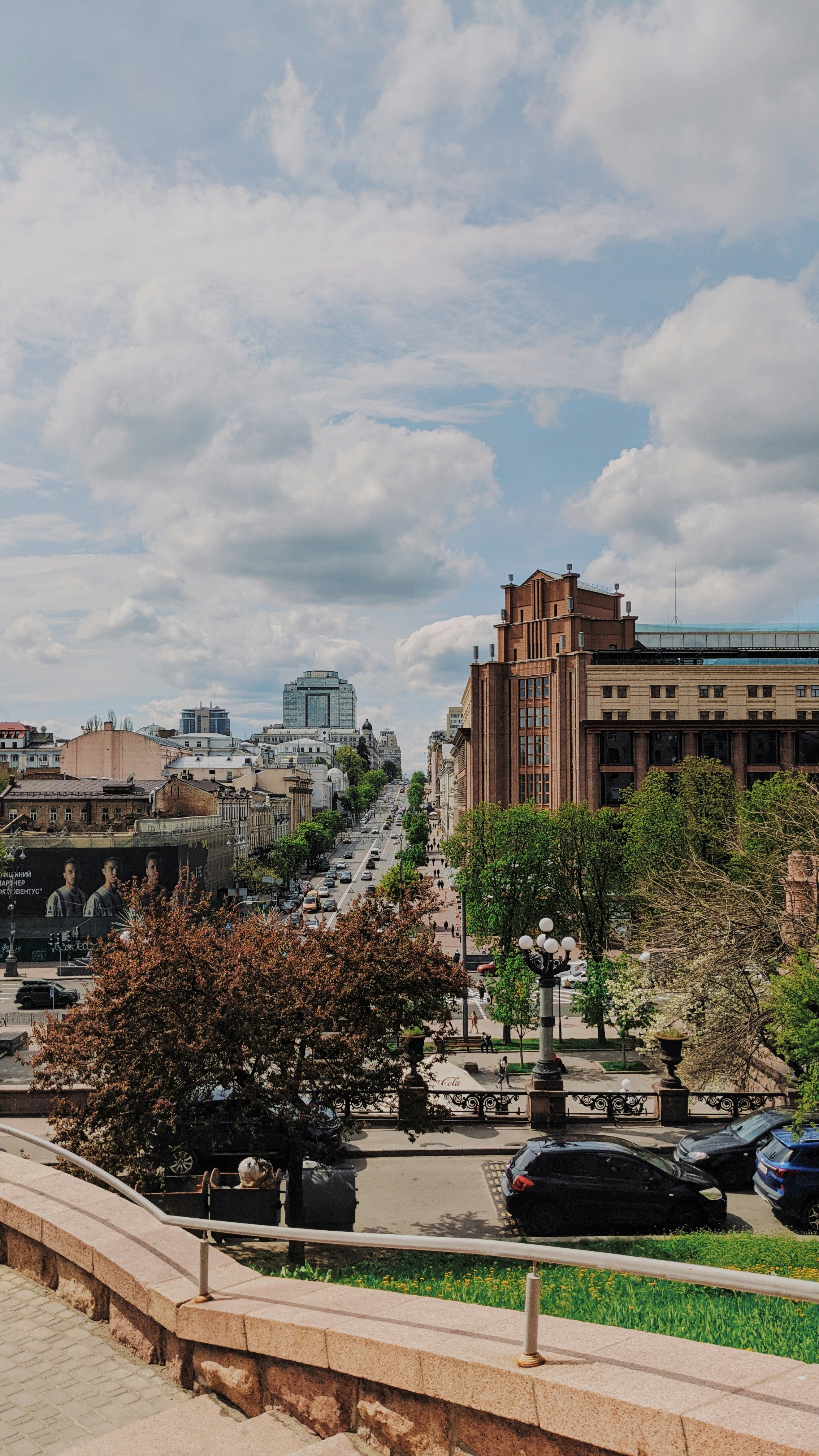 Vibrant cityscape showcasing a blend of modern and historical architecture along a bustling avenue, framed by lush greenery and dramatic clouds.