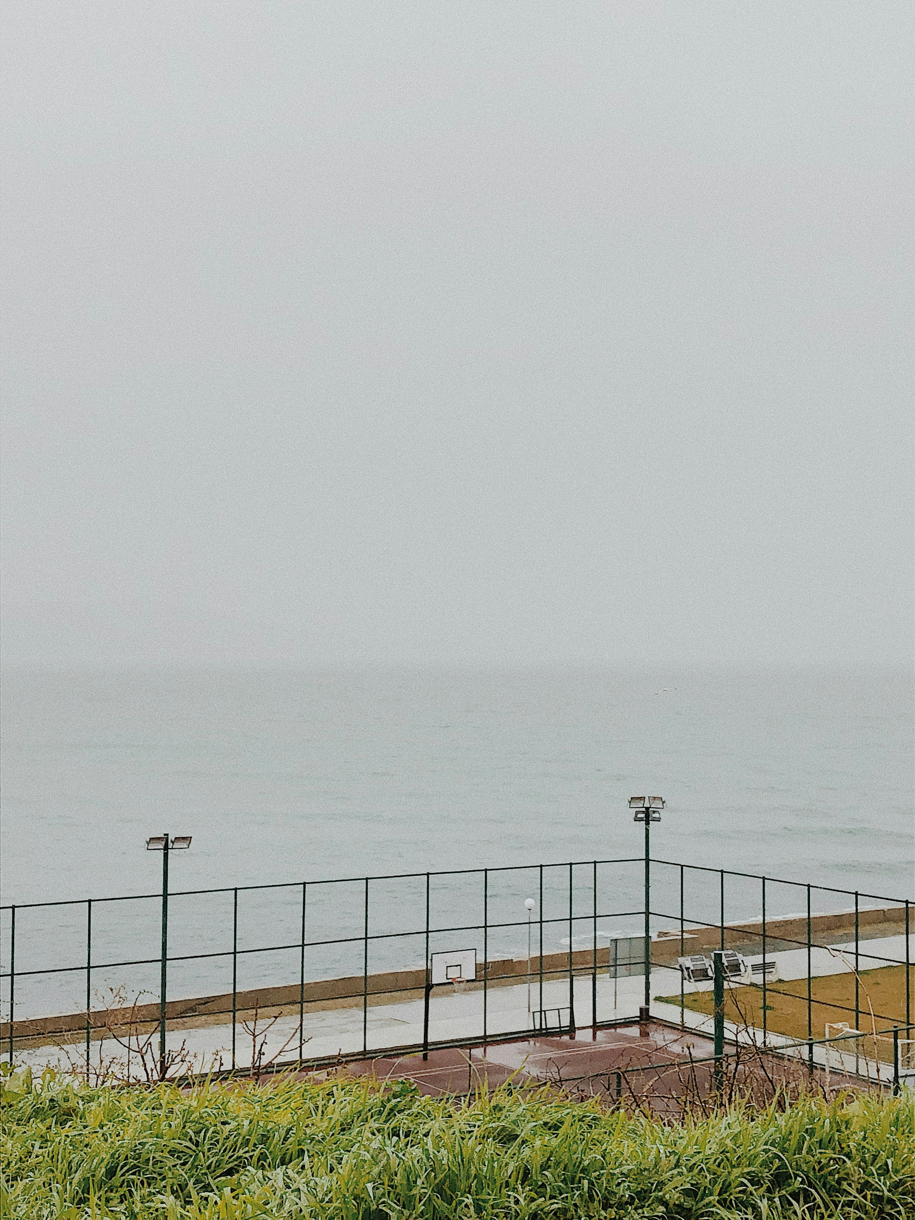 Basketball court overlooking a calm sea under a gray sky, with minimal human presence. The scene evokes a sense of tranquility and solitude.