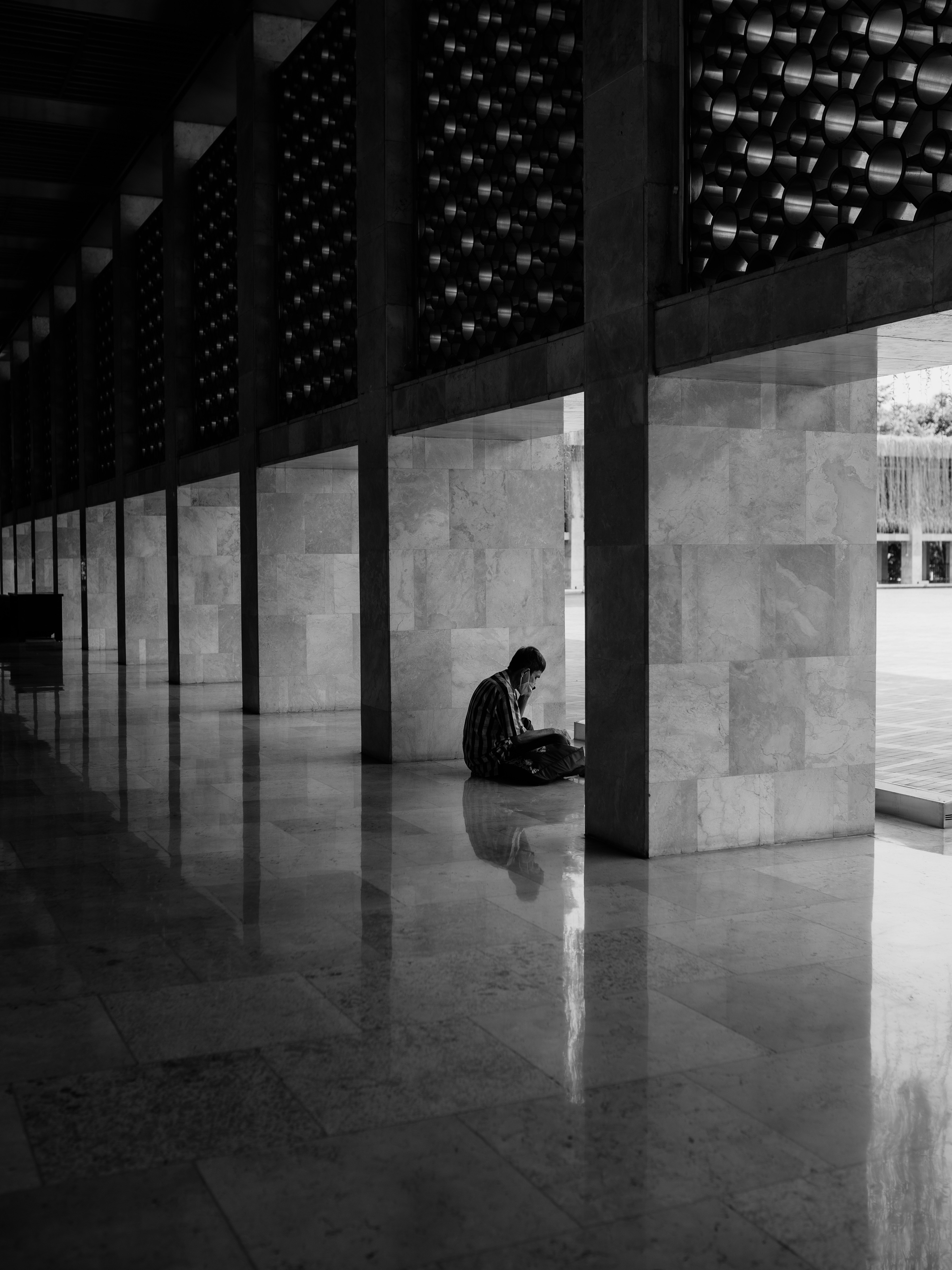 A solitary figure immersed in work, seated on polished marble floor beneath striking geometric architecture. The contrast of light and shadow enhances the serene atmosphere.