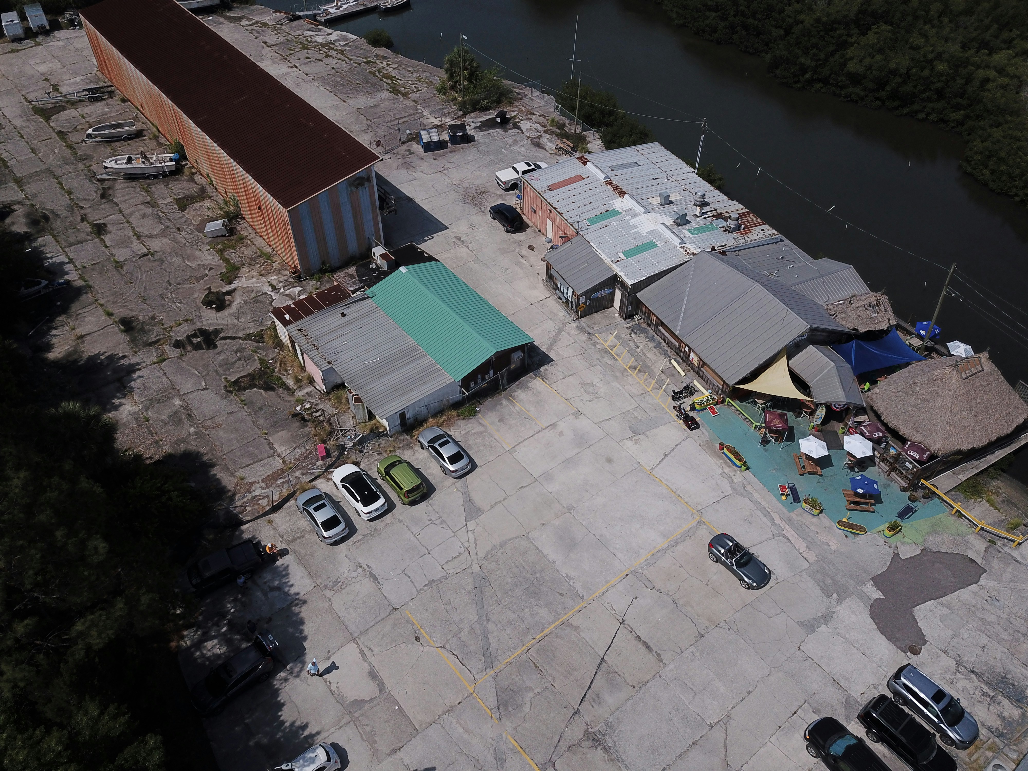 Aerial view of a rustic riverside venue featuring various structures, parked vehicles, and outdoor seating areas along the water's edge.