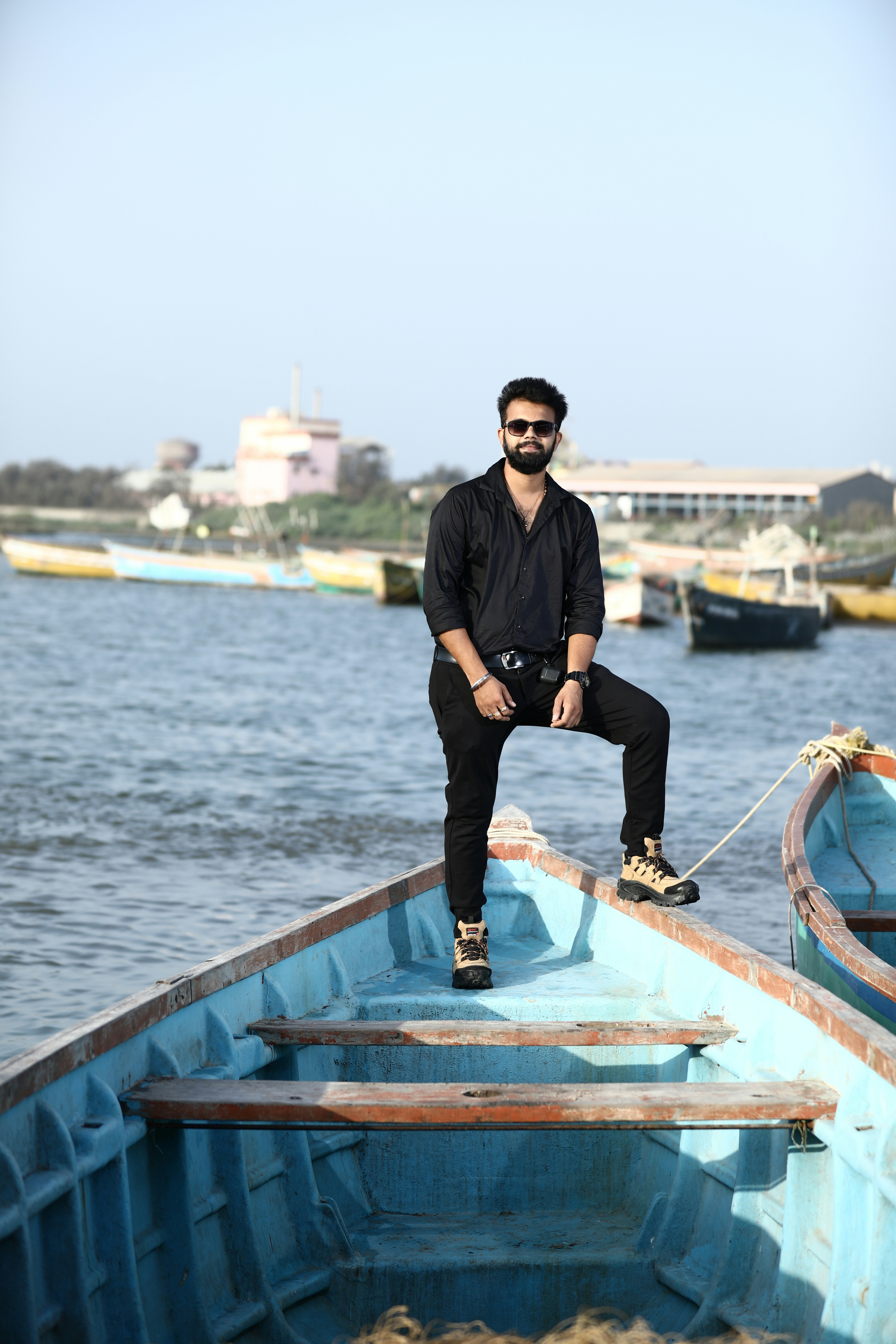A man in a black outfit confidently stands on the bow of a blue boat, overlooking a tranquil river with colorful fishing boats in the background.